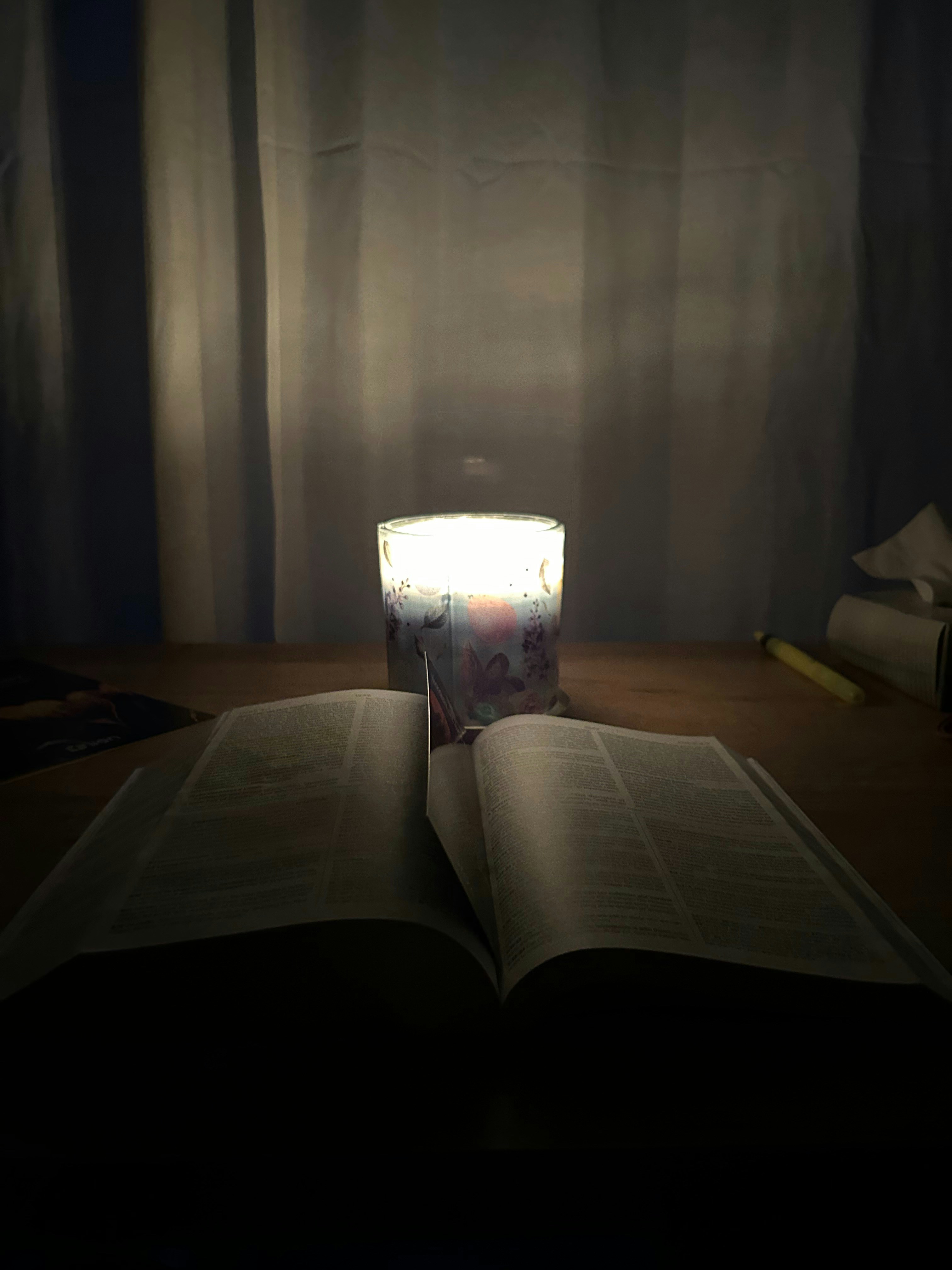 Candle on a wooden desk lights an open two-column book, casting soft shadows across the pages.