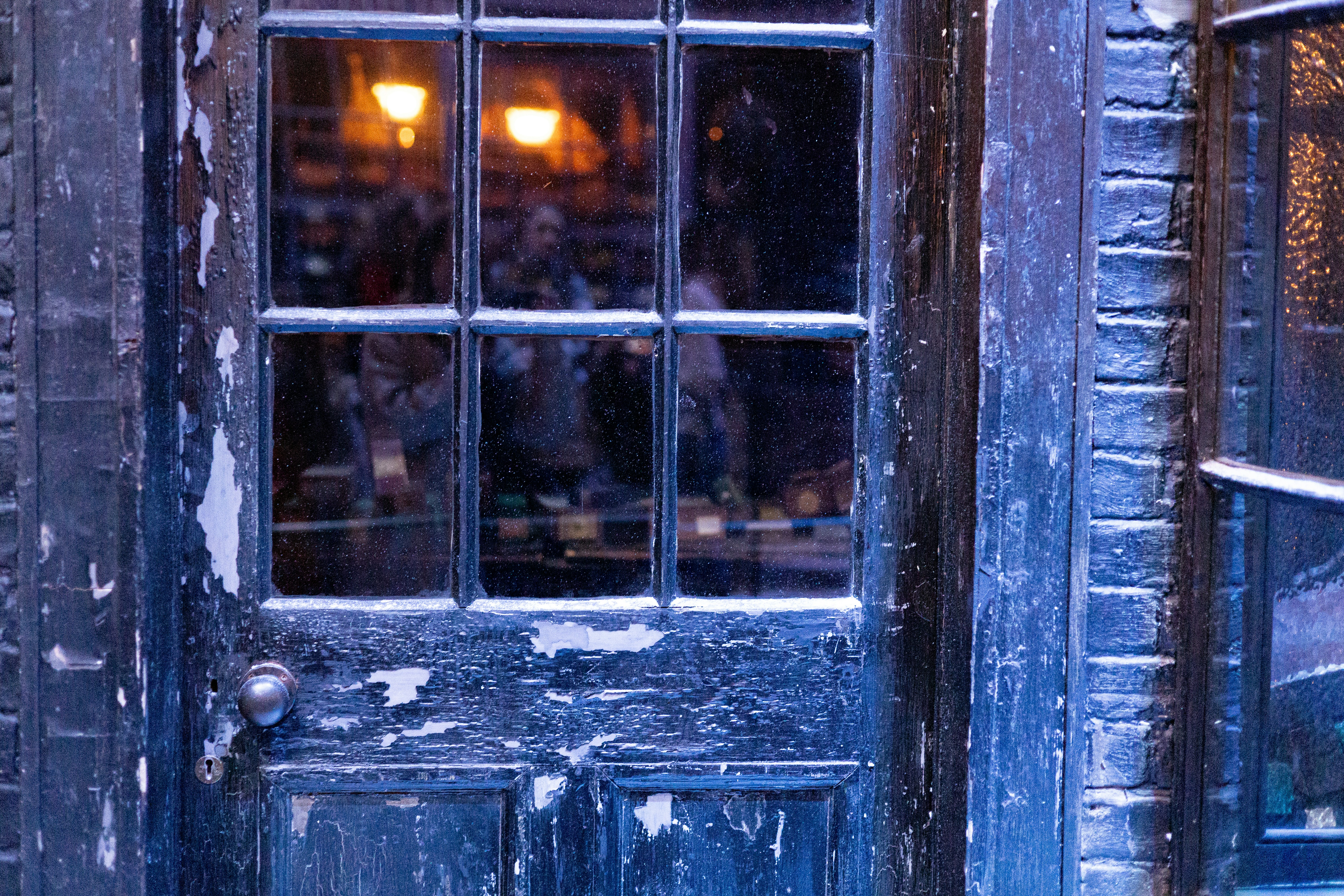 A rustic blue wooden door with peeling paint, set against a brick wall, reflecting the warm glow of interior lights.
