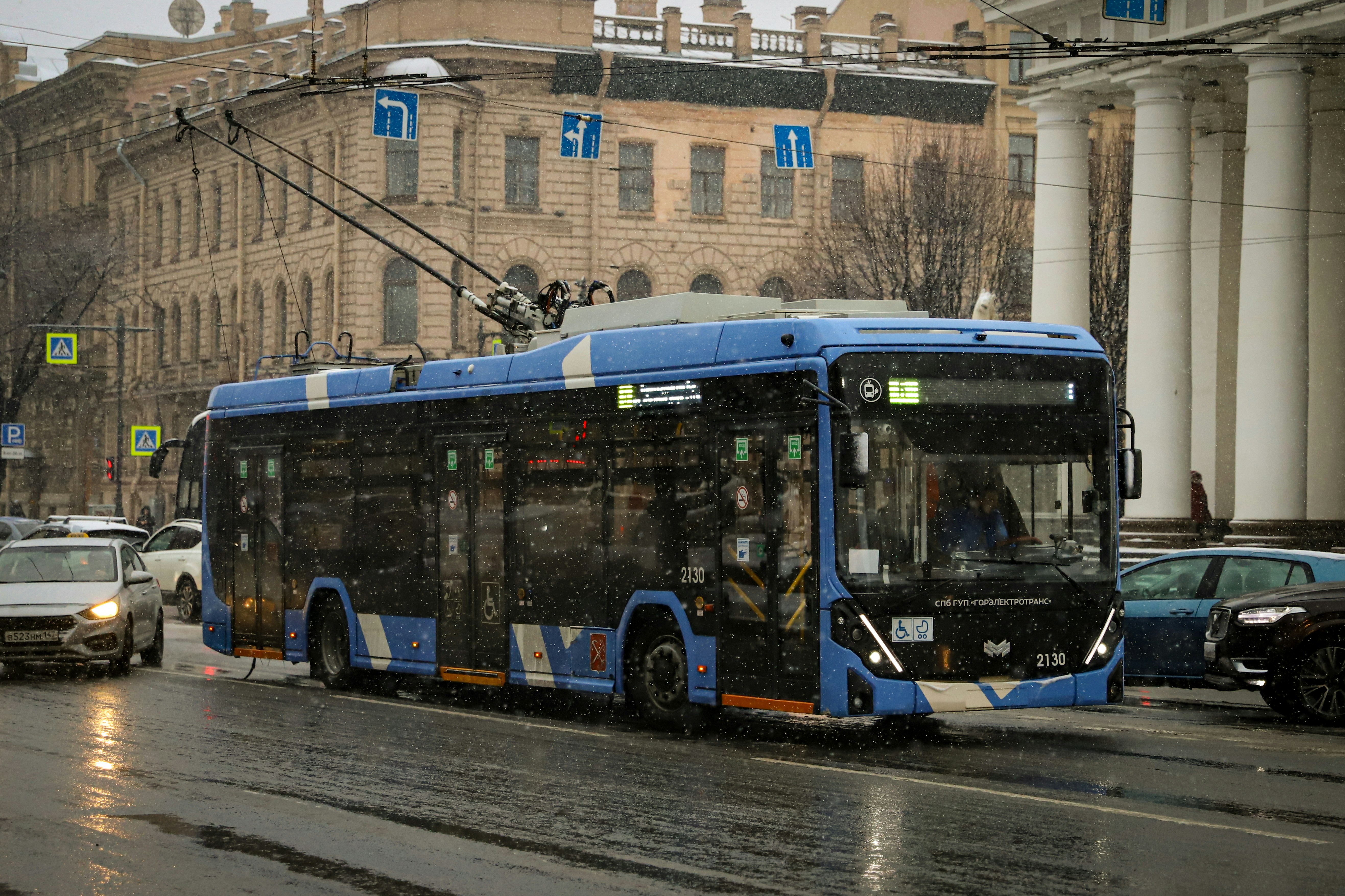 A blue and white trolleybus navigates a snowy city street, surrounded by historic architecture and falling snowflakes.