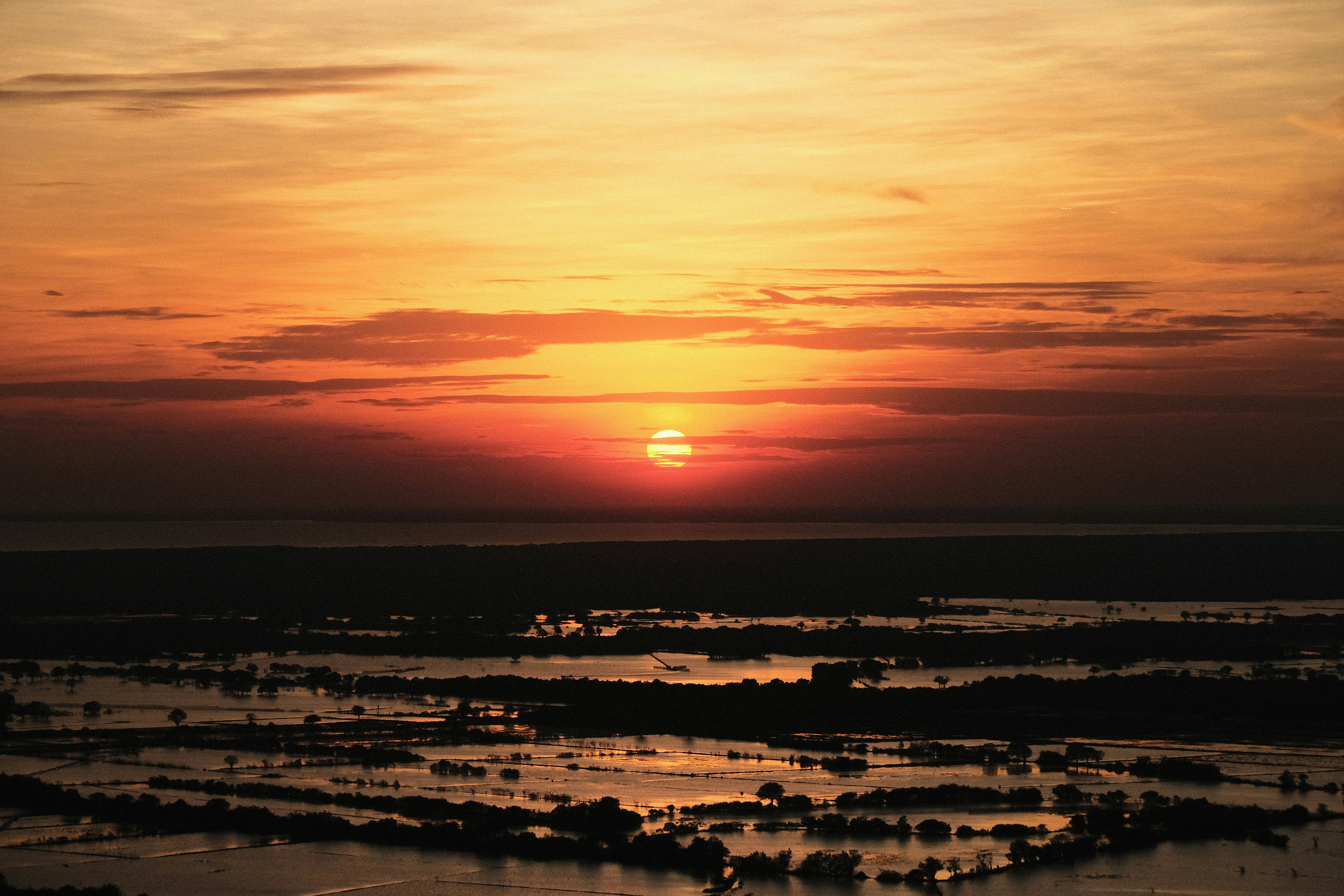 Sunset casting warm hues over expansive flooded fields with scattered silhouettes of trees.