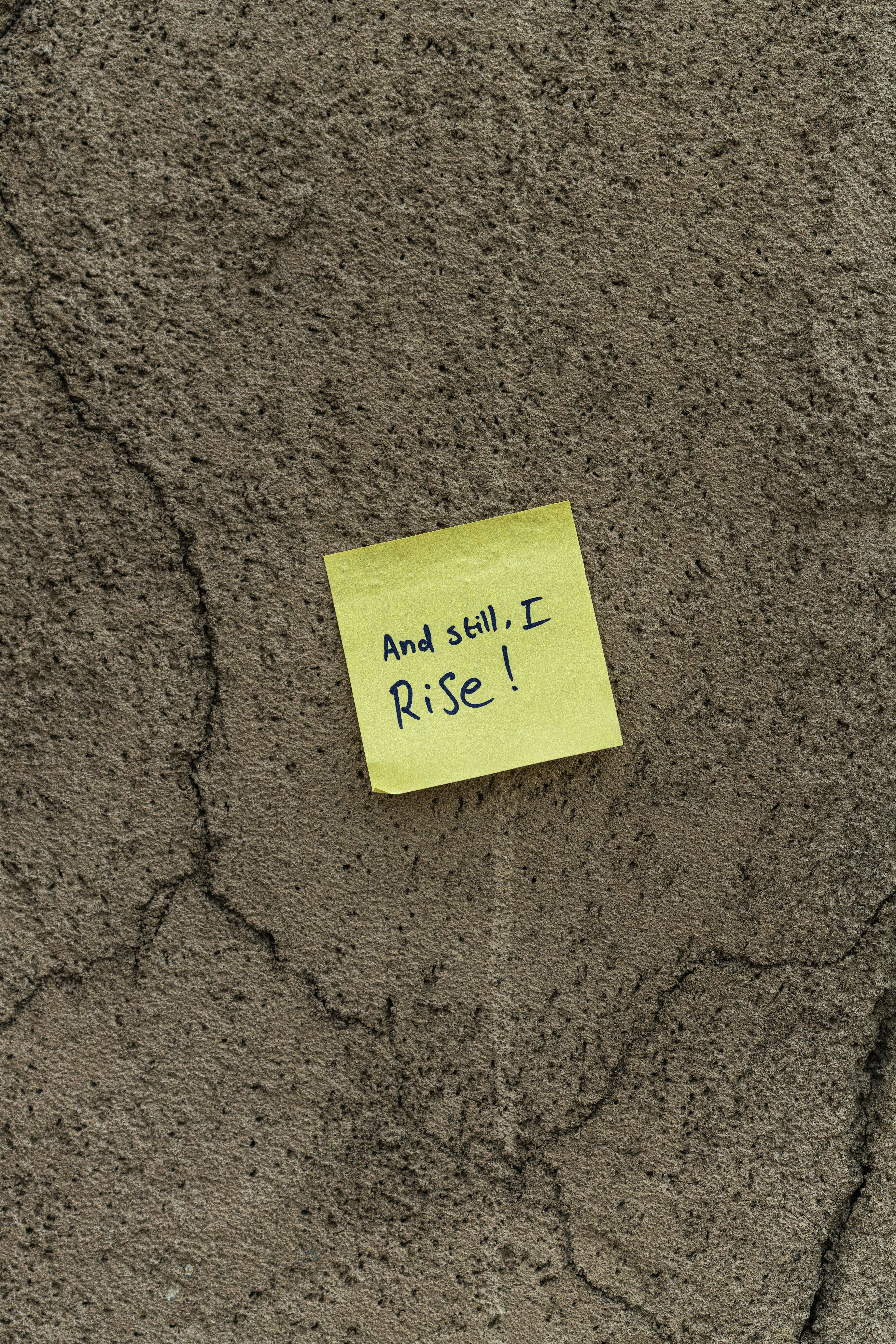 A yellow sticky note bearing 'And still, I Rise!' rests on rough cracked earth, photographed in a close, textured shot.
