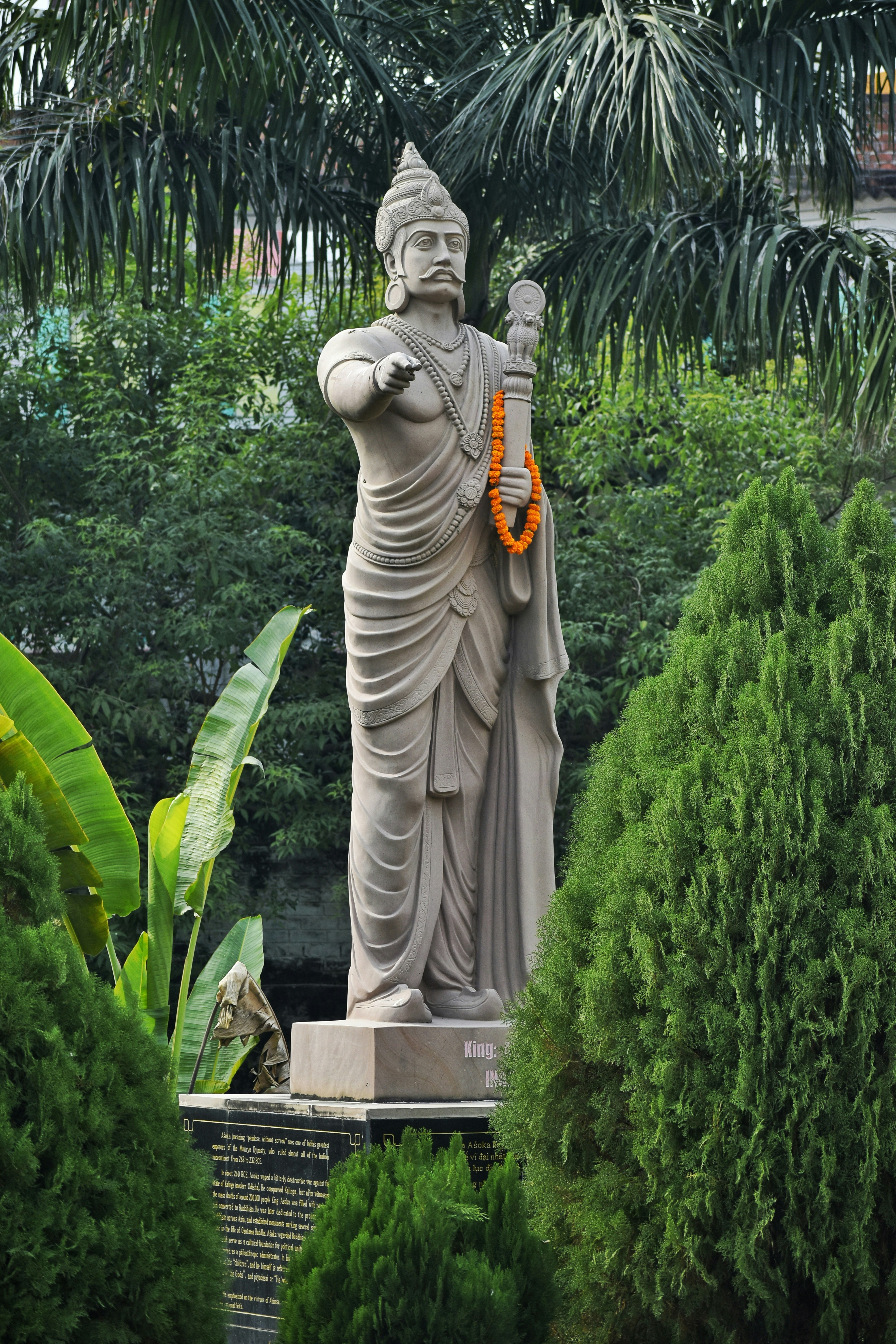 A detailed statue of a deity adorned with an orange garland, standing amidst lush greenery and plants.