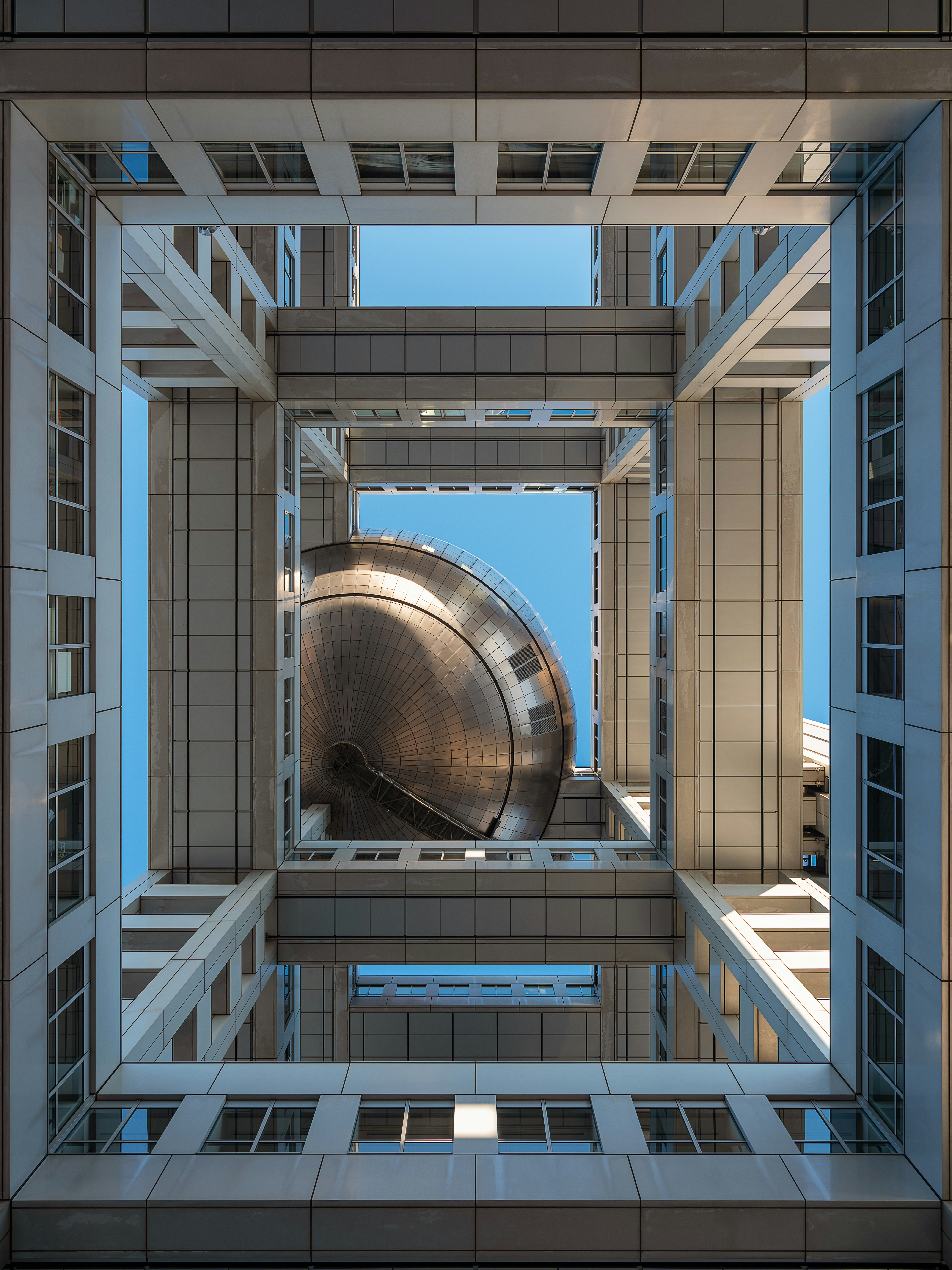 Architectural courtyard framed by a square grid of white corridors converging toward a central circular metallic core against a clear blue sky. Photograph.