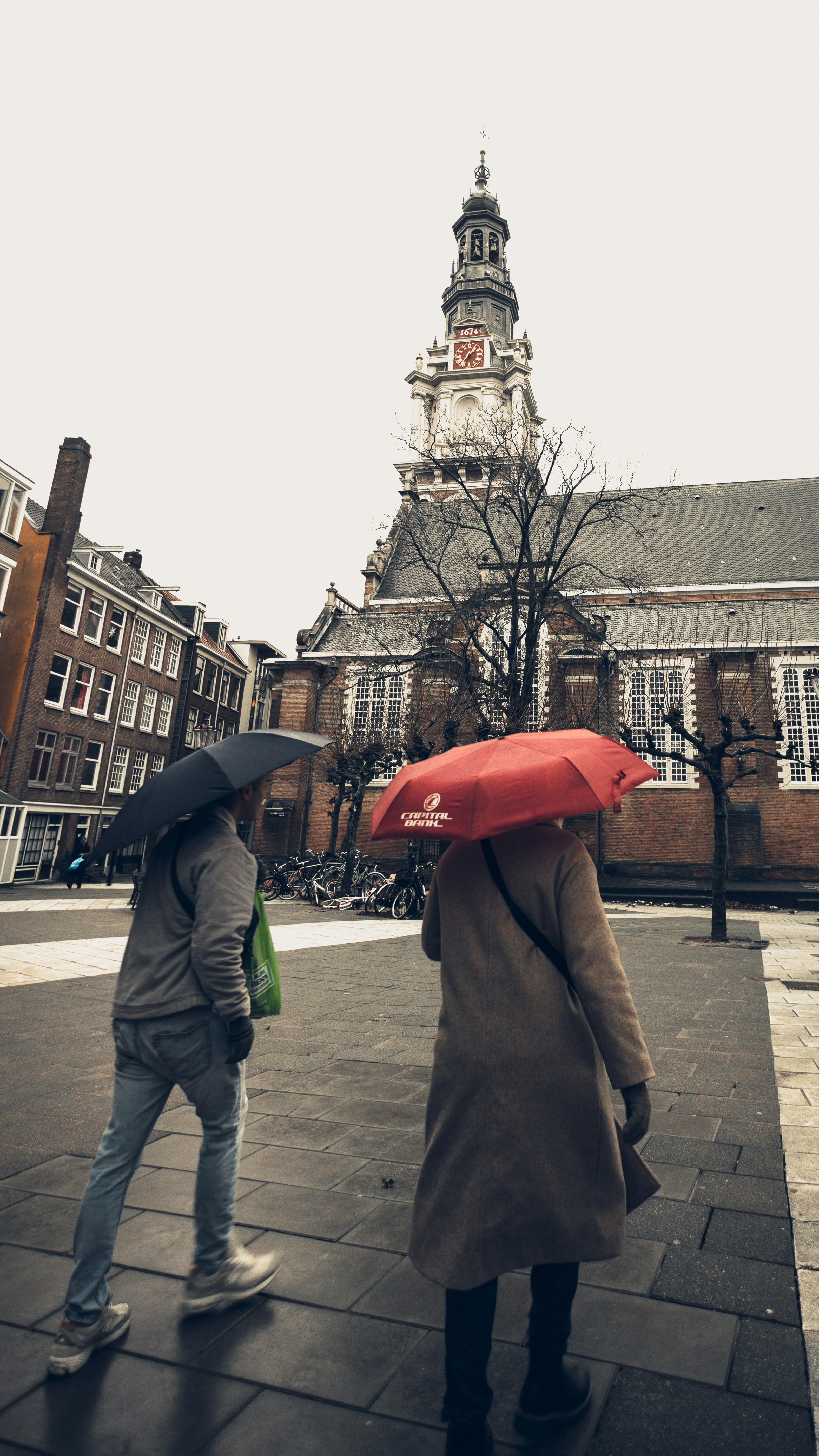This photograph captures a grey, overcast day with two pedestrians walking on the cobbled streets, each holding an umbrella against the drizzle. In the background stands a historic building with a prominent clock tower, suggesting a location in a European city. The bare trees and the architectural style give a sense of the season and the region's cultural heritage.