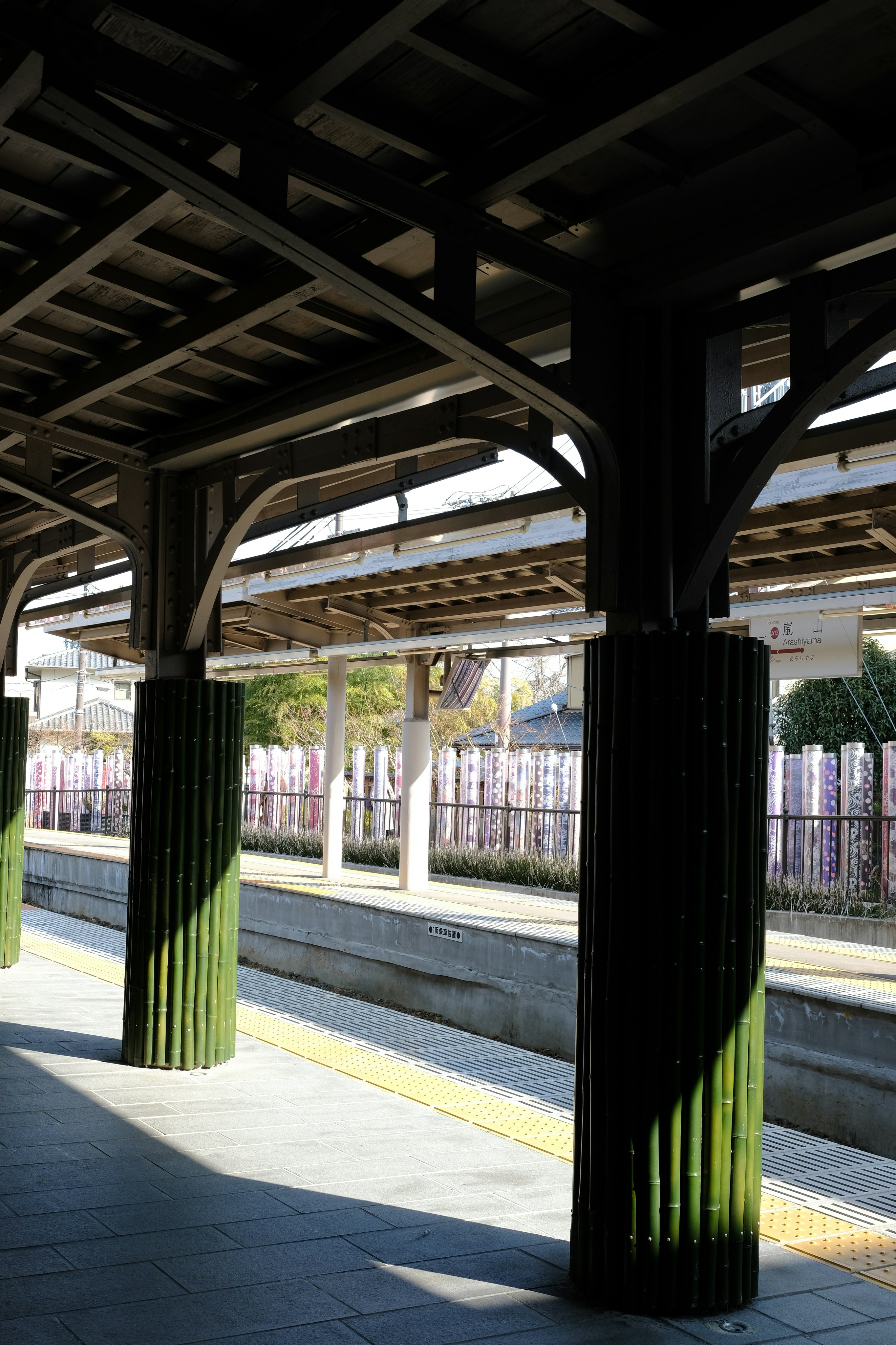 Sunlight casts geometric shadows on bamboo-clad columns at a train platform, highlighting architectural details.