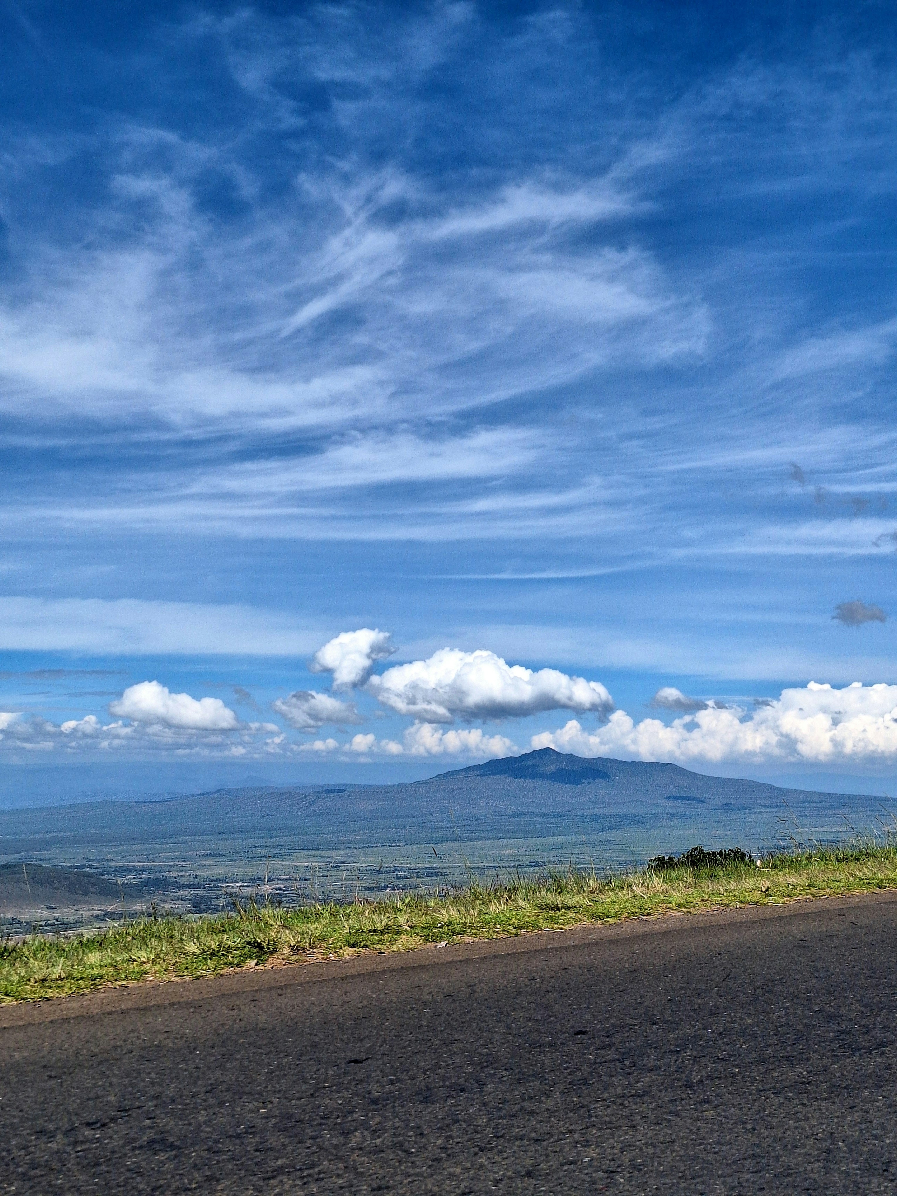 A landscape photograph of a roadside viewpoint with a distant ridge under a bright blue sky and scattered clouds.