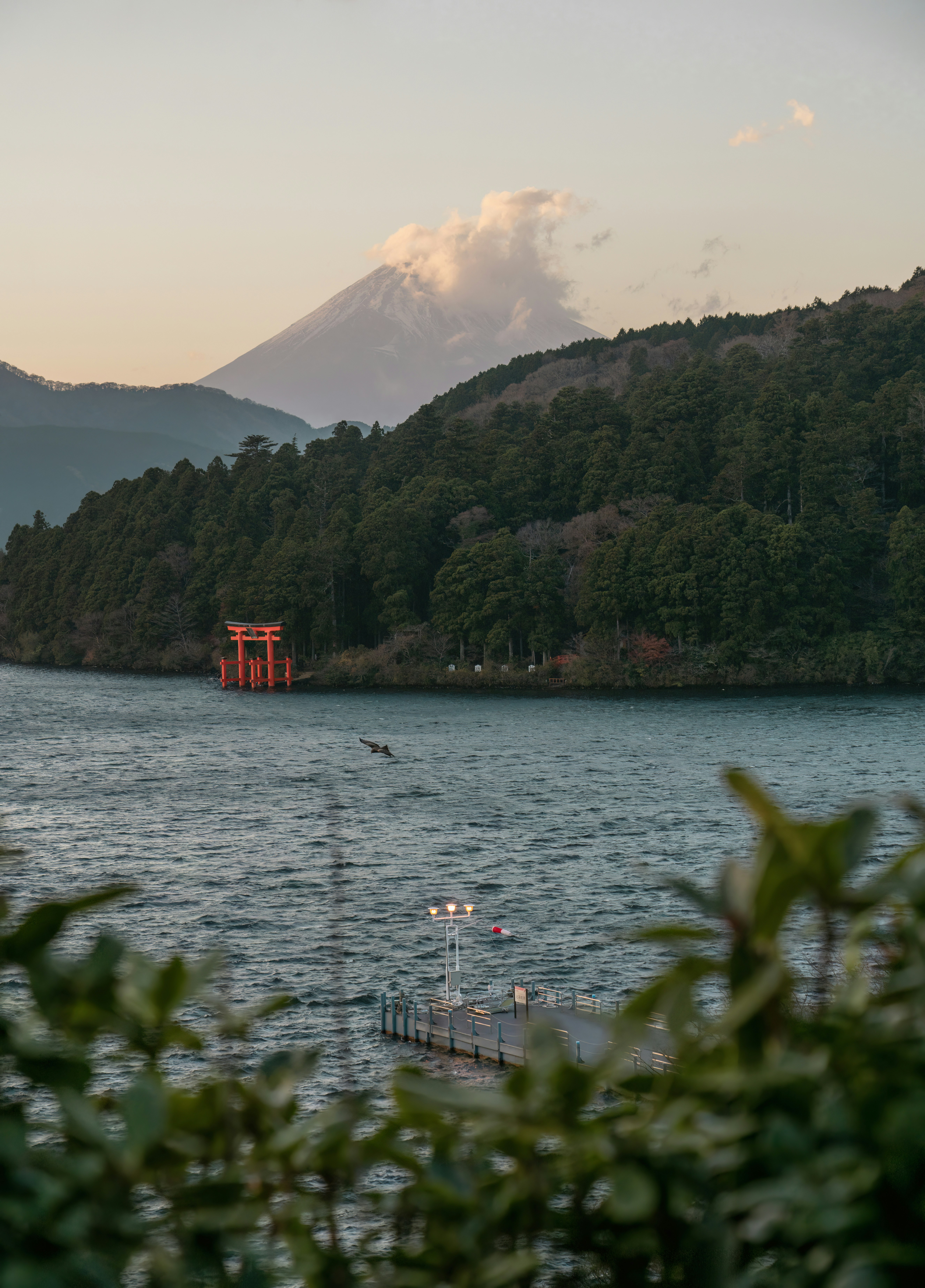 Torii gate stands by the tranquil waters with Mount Fuji in the background, framed by lush greenery. A serene moment captured at dusk.