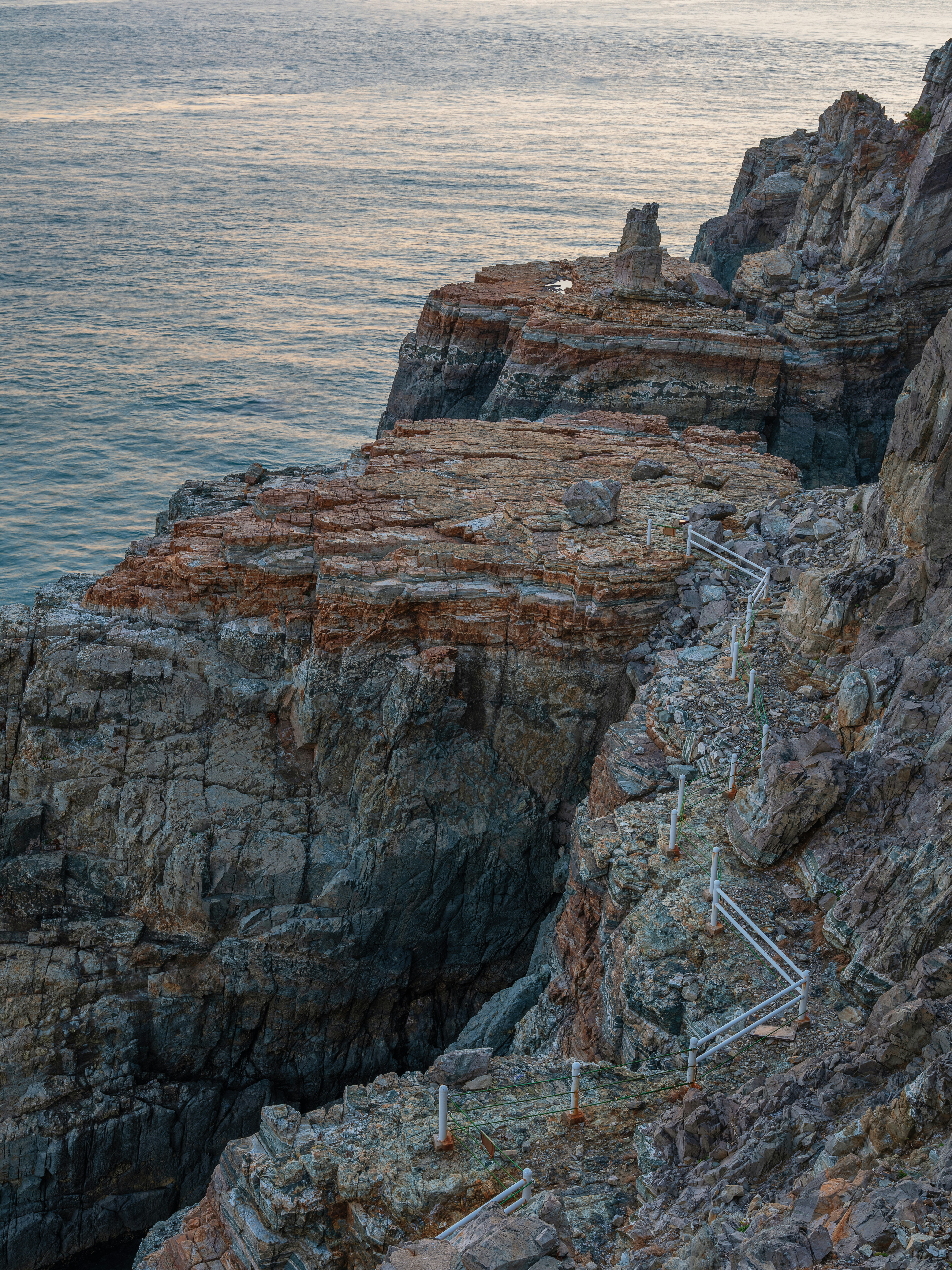 Weathered cliff face features a zigzag metal railing staircase descending toward the sea, lit by warm dusk light.