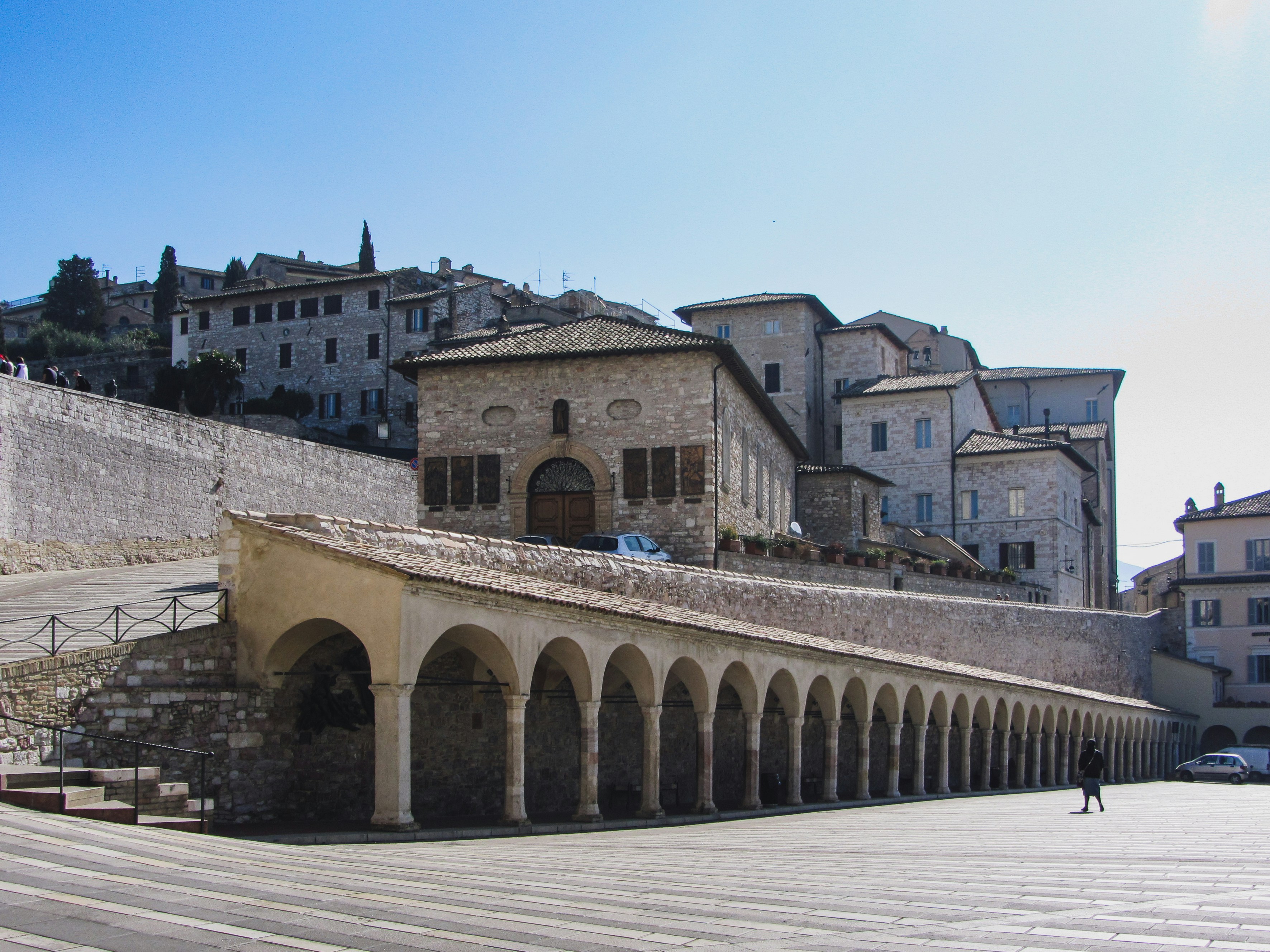 Historic stone arches line a sunlit square with distant hillside buildings.