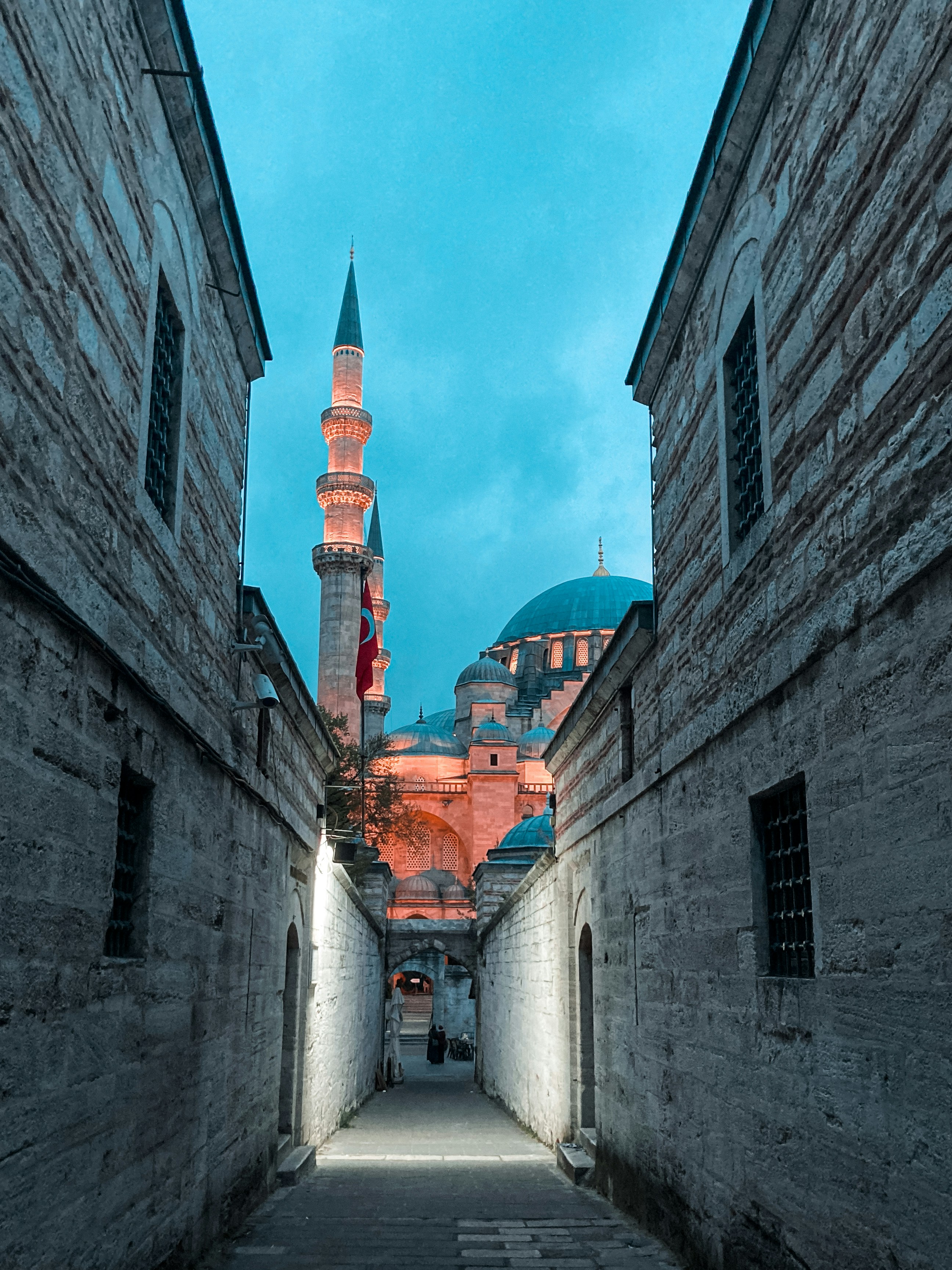 Narrow alleyway framed by ancient stone walls leading to a mosque, illuminated by soft evening light.