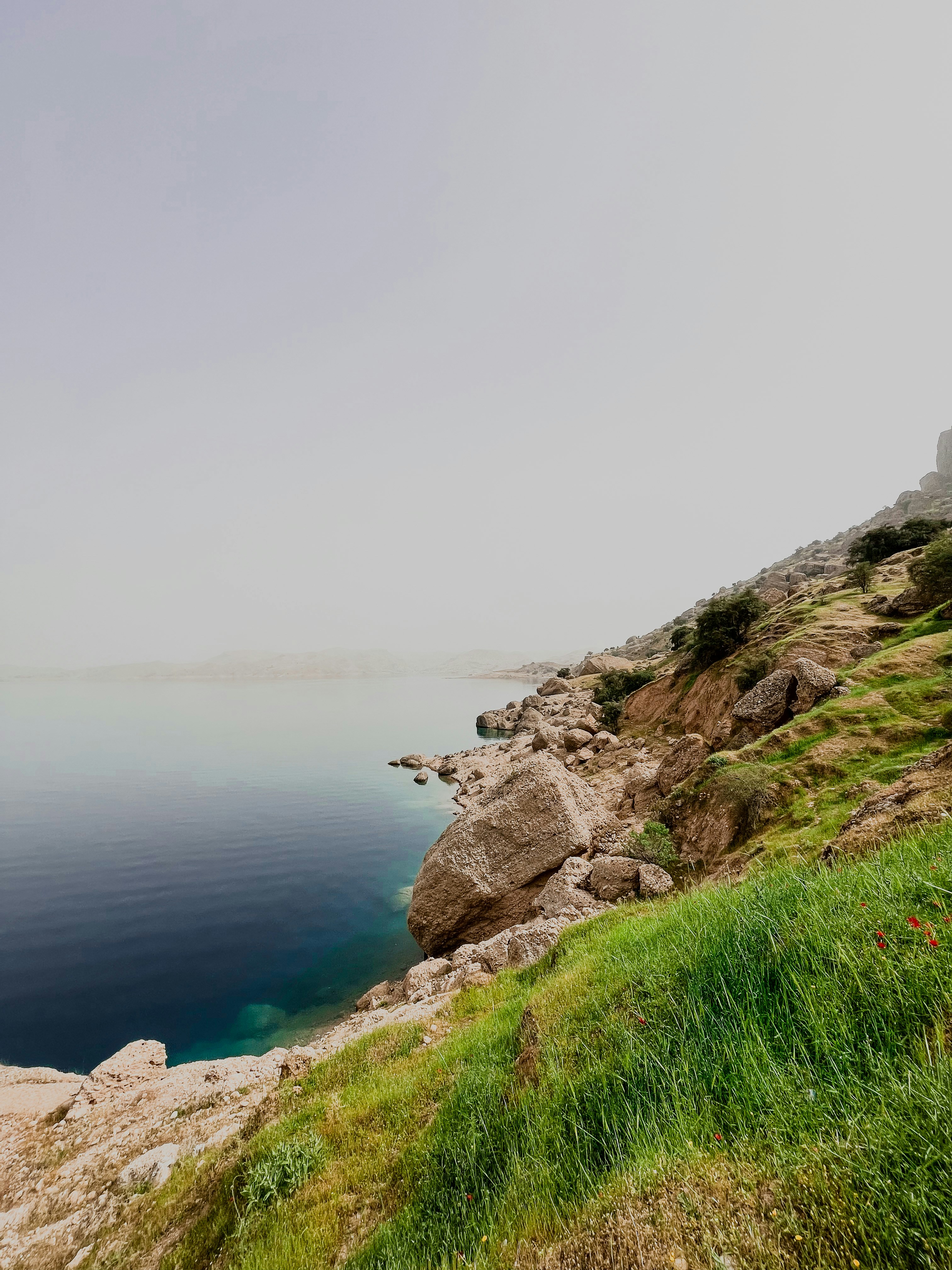 Tranquil shoreline with rocky formations and lush greenery, reflecting calm waters under a hazy sky.