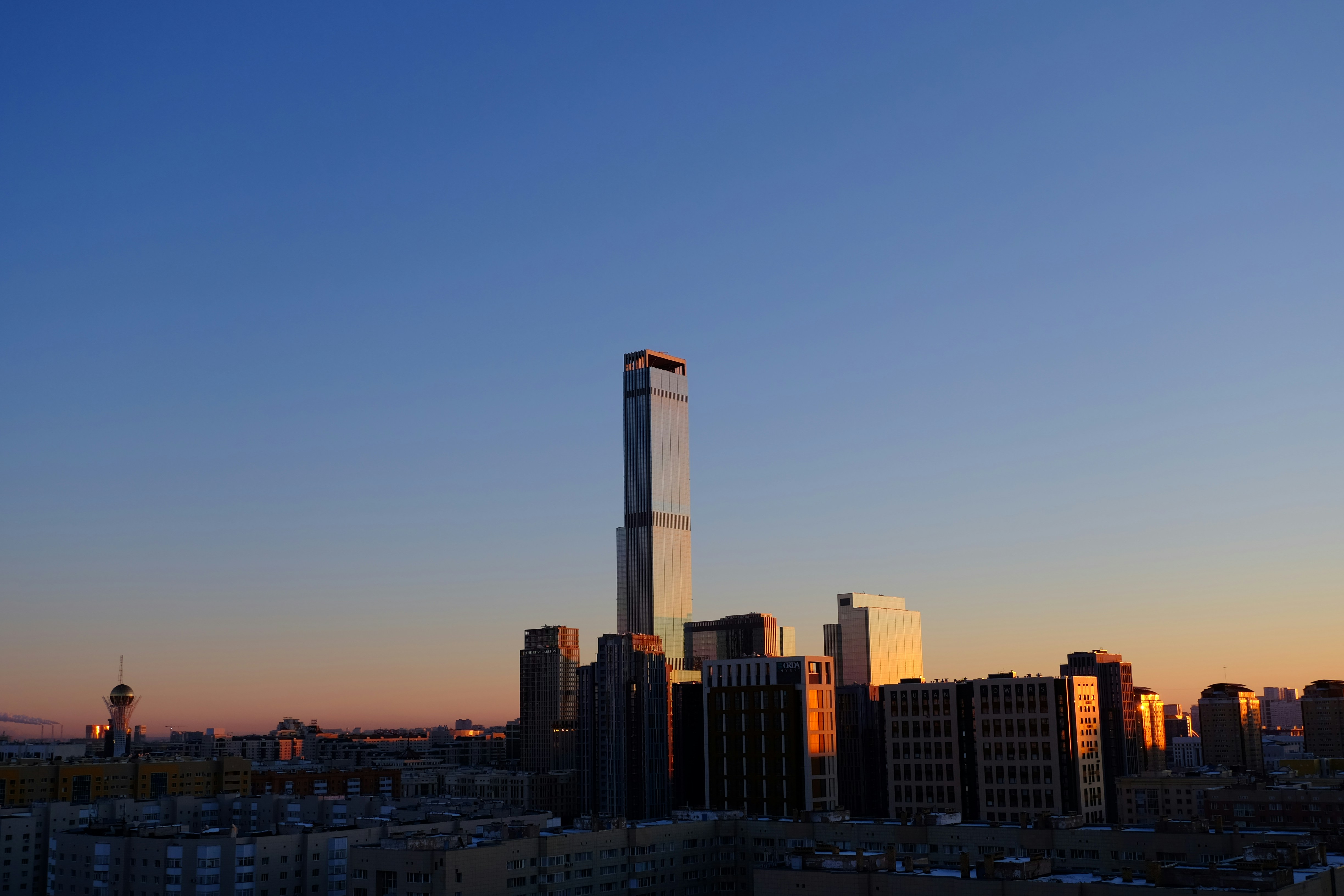 City skyline illuminated by the warm hues of sunset, featuring a prominent skyscraper and surrounding buildings. 