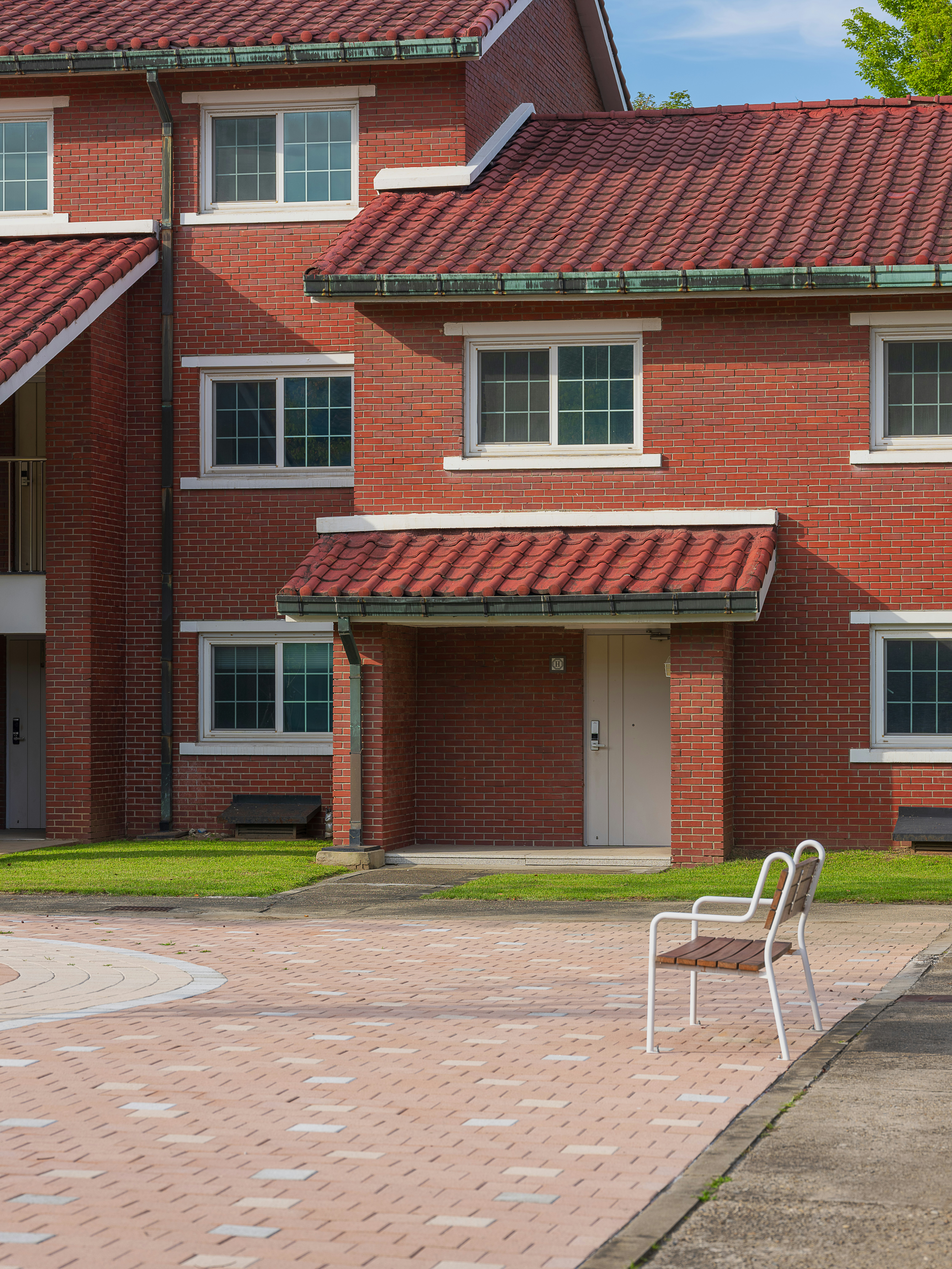 Photograph of a sunlit red-brick building with white-framed windows and a small porch, set in a brick-paved courtyard with a lone bench.