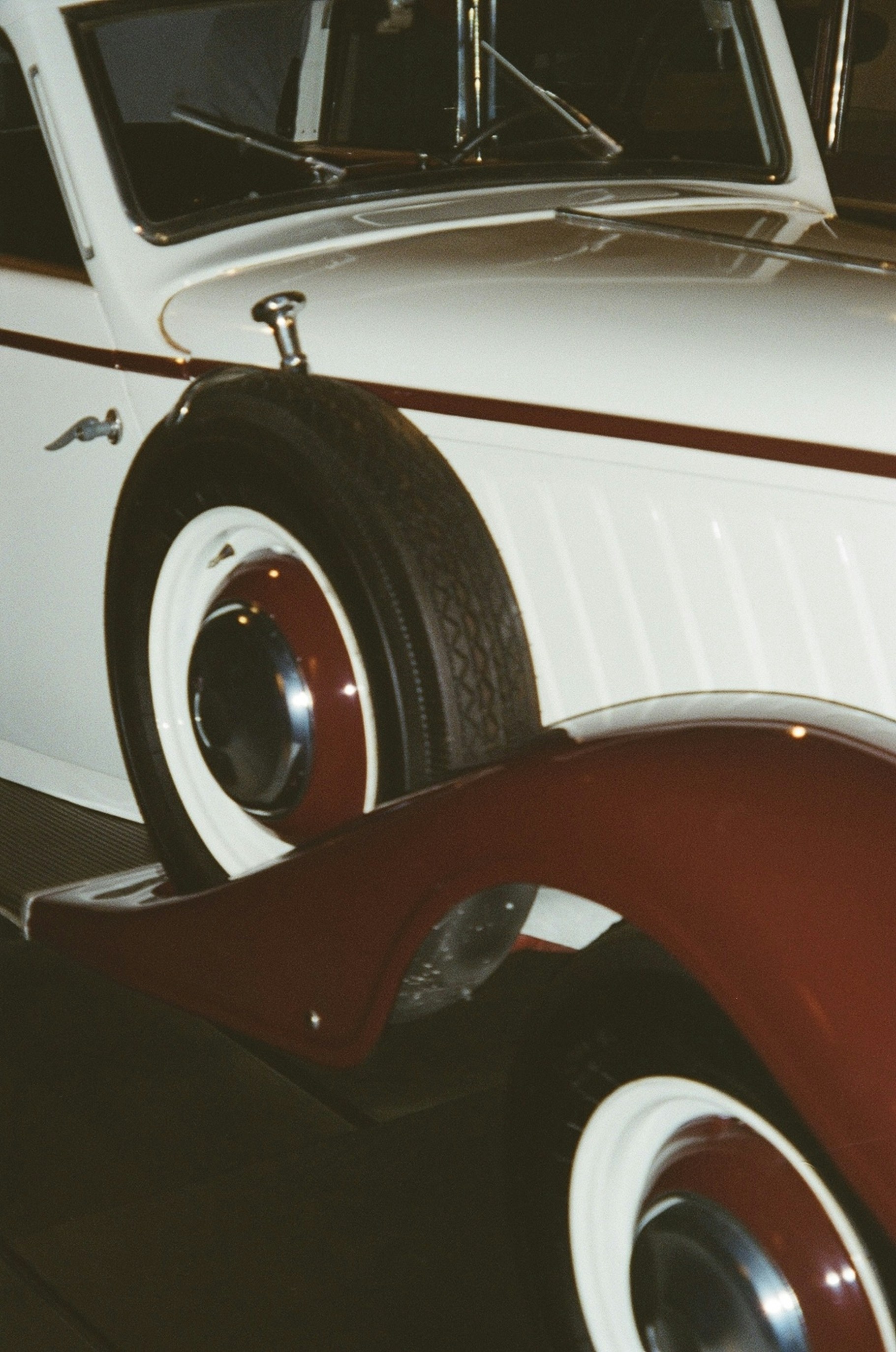 Close-up of a vintage white car front featuring a whitewall tire and chrome hubcap. Red fenders curve around the wheel in low light.
