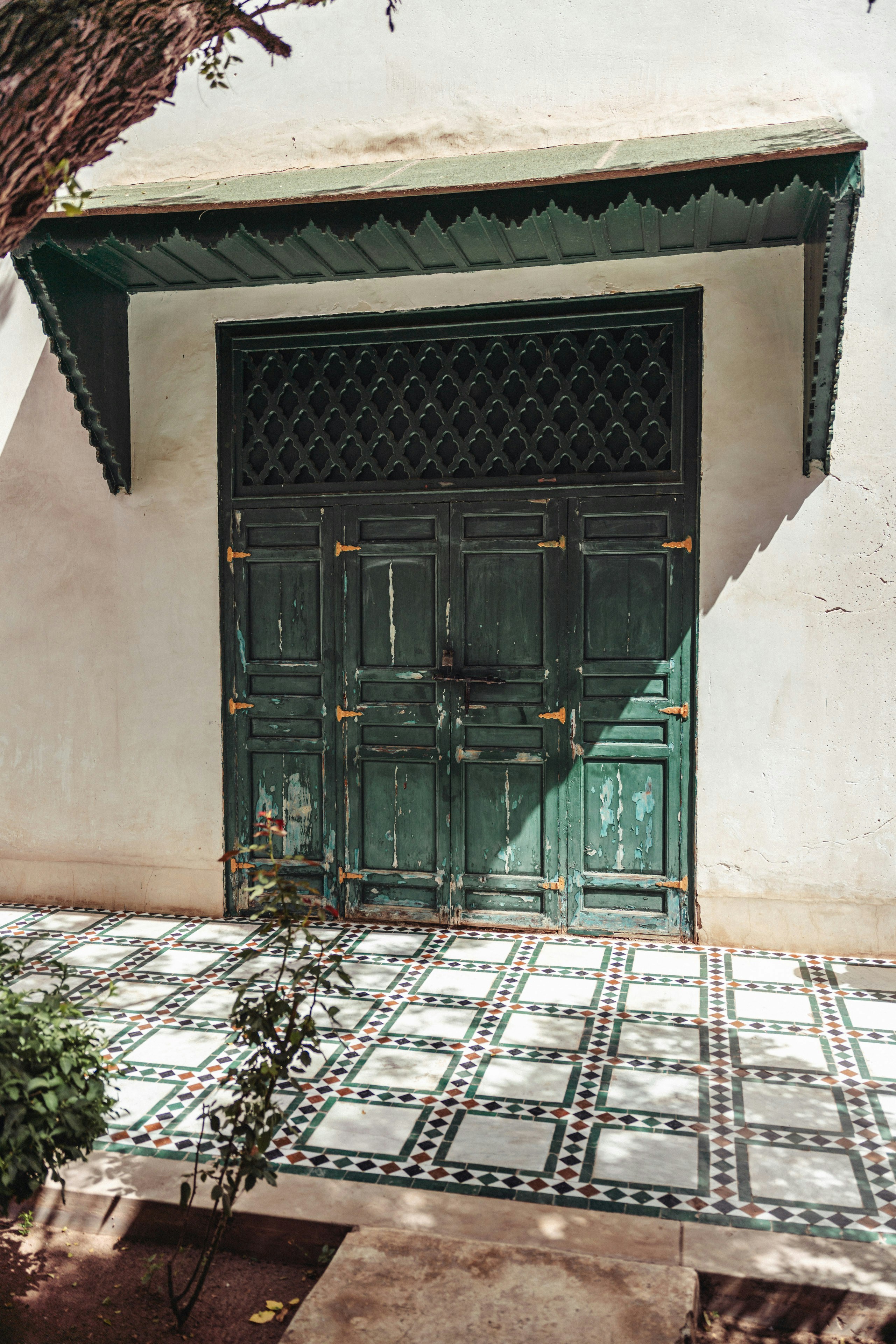 Photograph of an aged green wooden door with a lattice transom, set in a sunlit beige wall above a checkerboard tile courtyard.