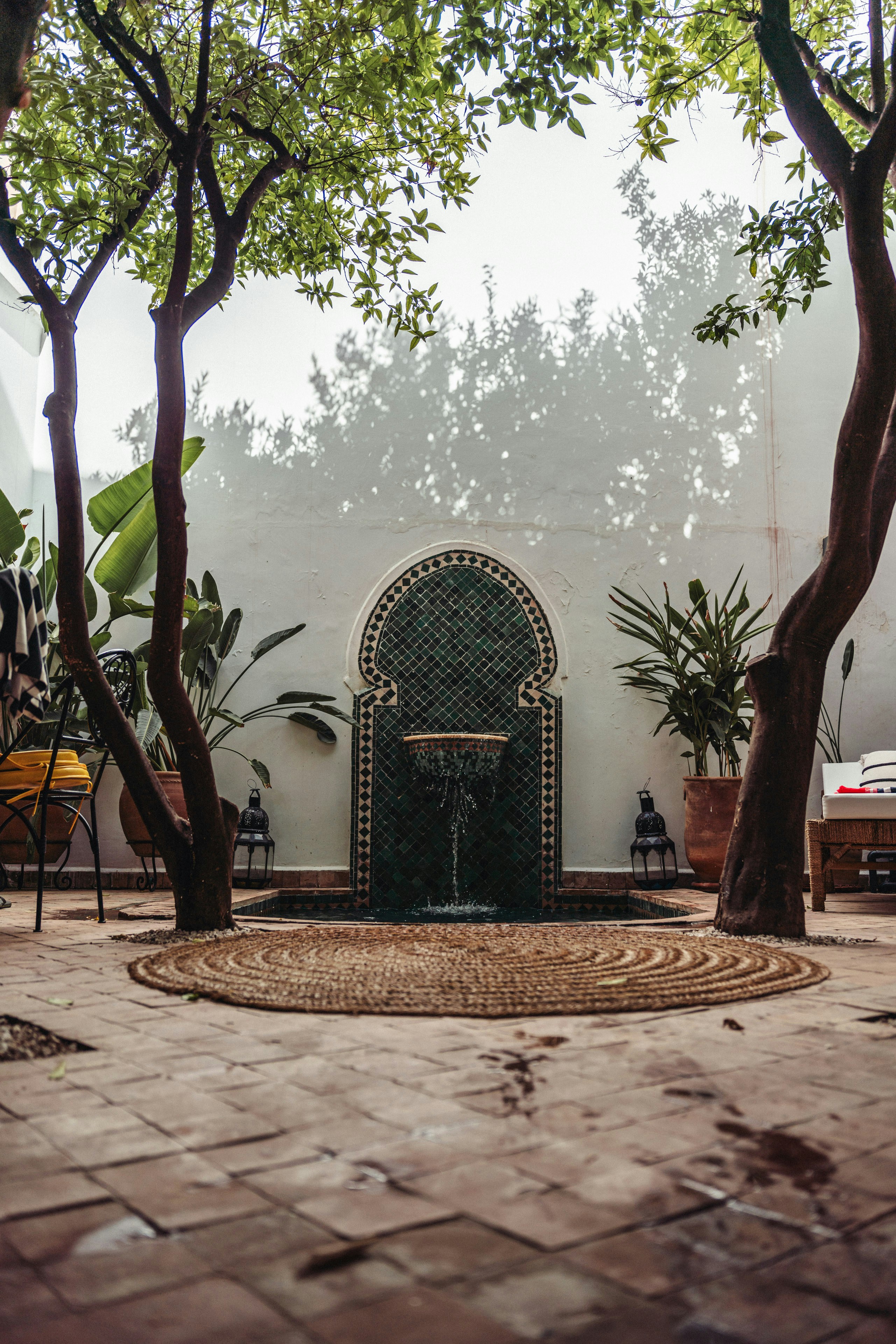 Central arched tile fountain in a sunlit courtyard, framed by trees and potted plants.