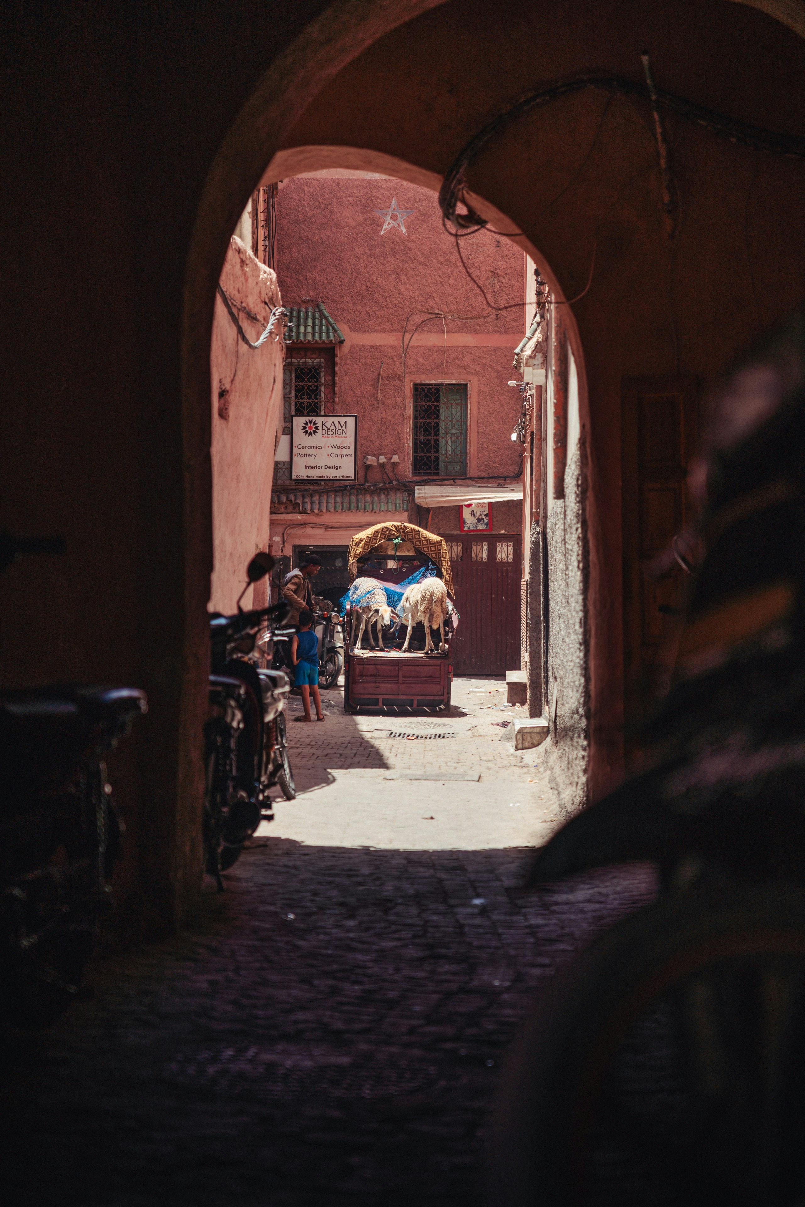 A photograph of a narrow alley framed by an arch, showing a market cart with oxen under a blue canopy and pedestrians in the sunlit background.