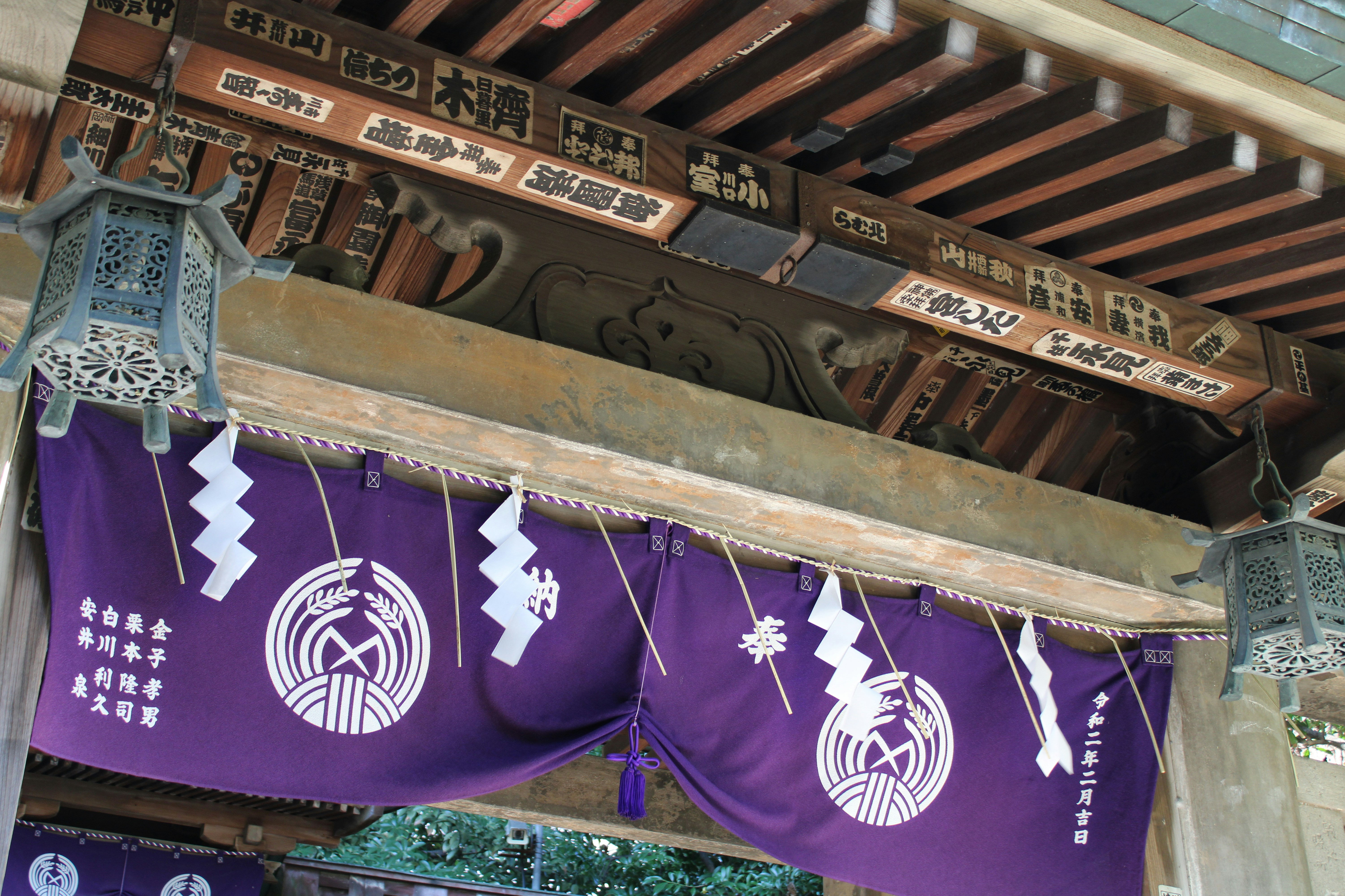 Purple ceremonial banners with white symbols hang from a wooden shrine adorned with intricate carvings and lanterns.
