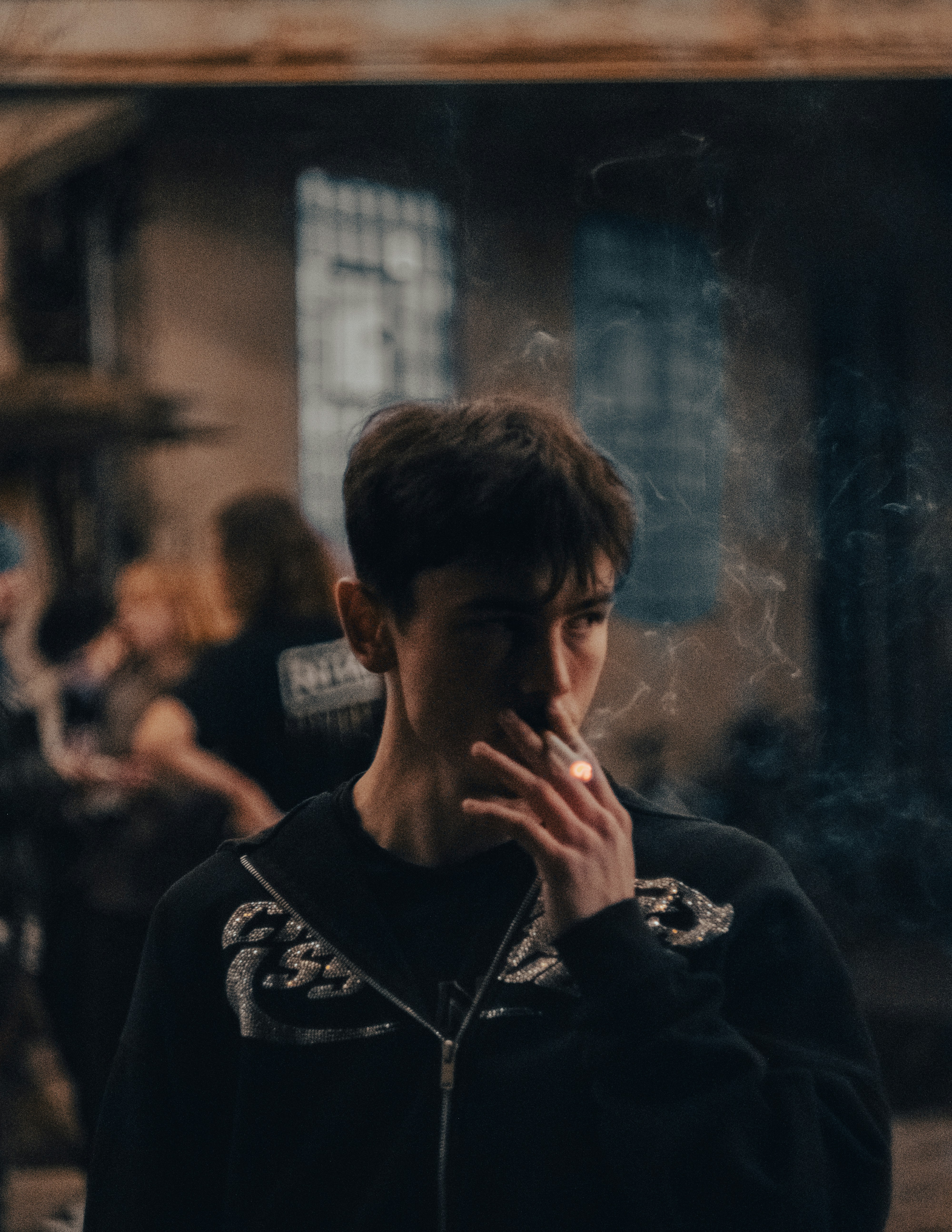 Young man smoking in a dim, crowded room with warm lighting and hazy reflections.