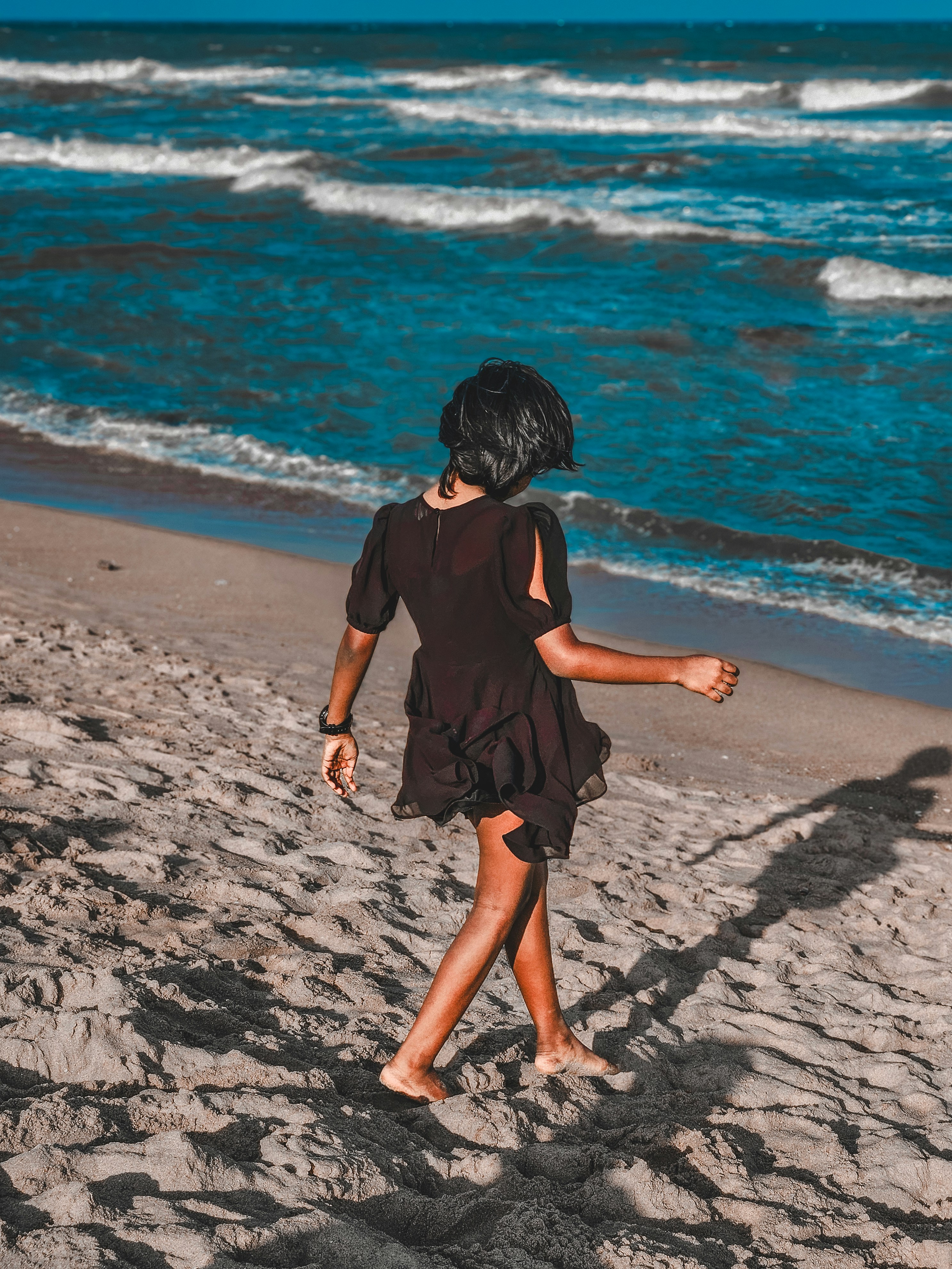 A figure in a flowing black dress dances along the sandy beach, with gentle waves lapping at the shore. Sunlight casts playful shadows on the sand.