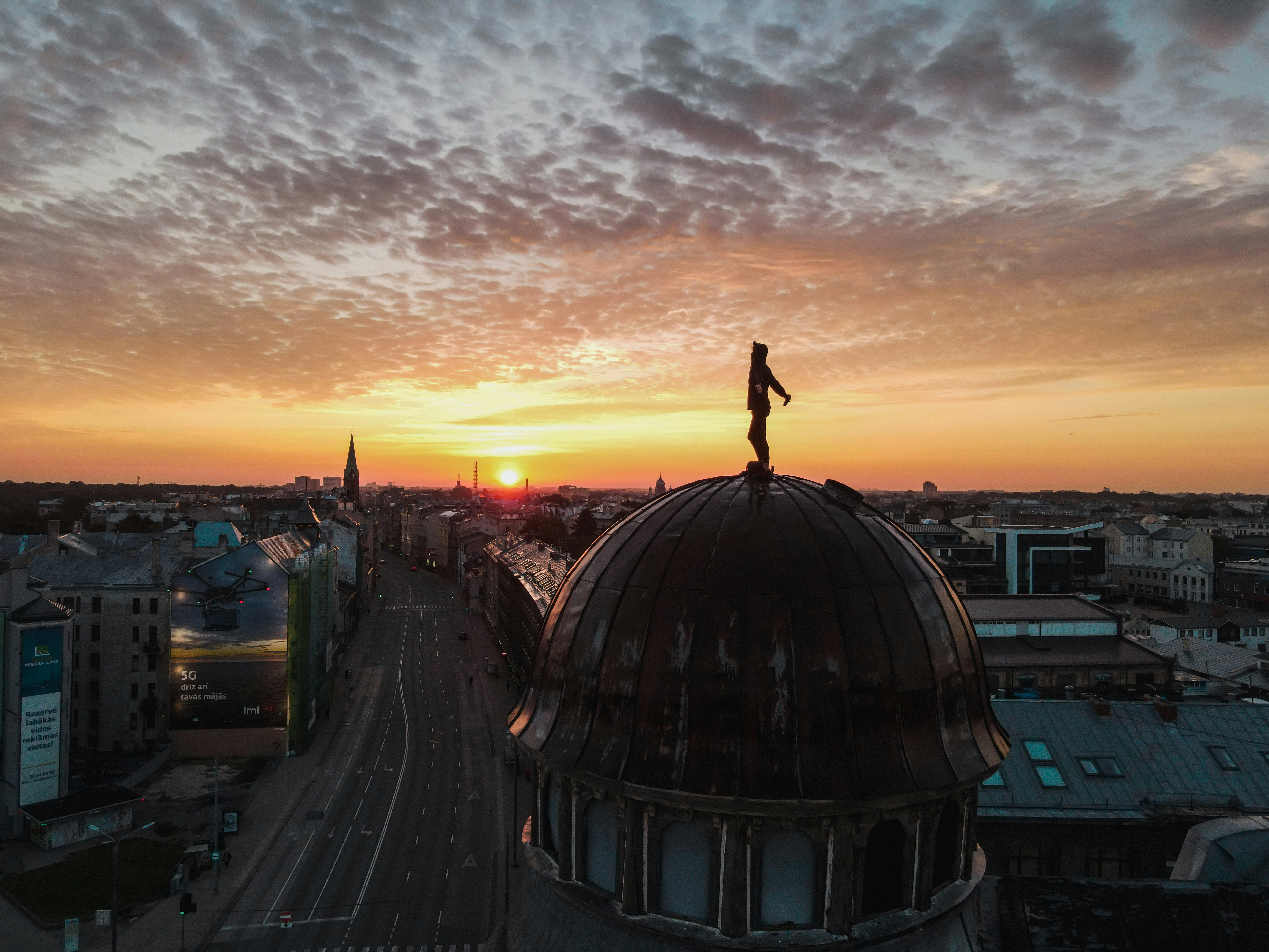 Silhouette of a figure atop a dome against a vibrant sunrise sky in a quiet cityscape.