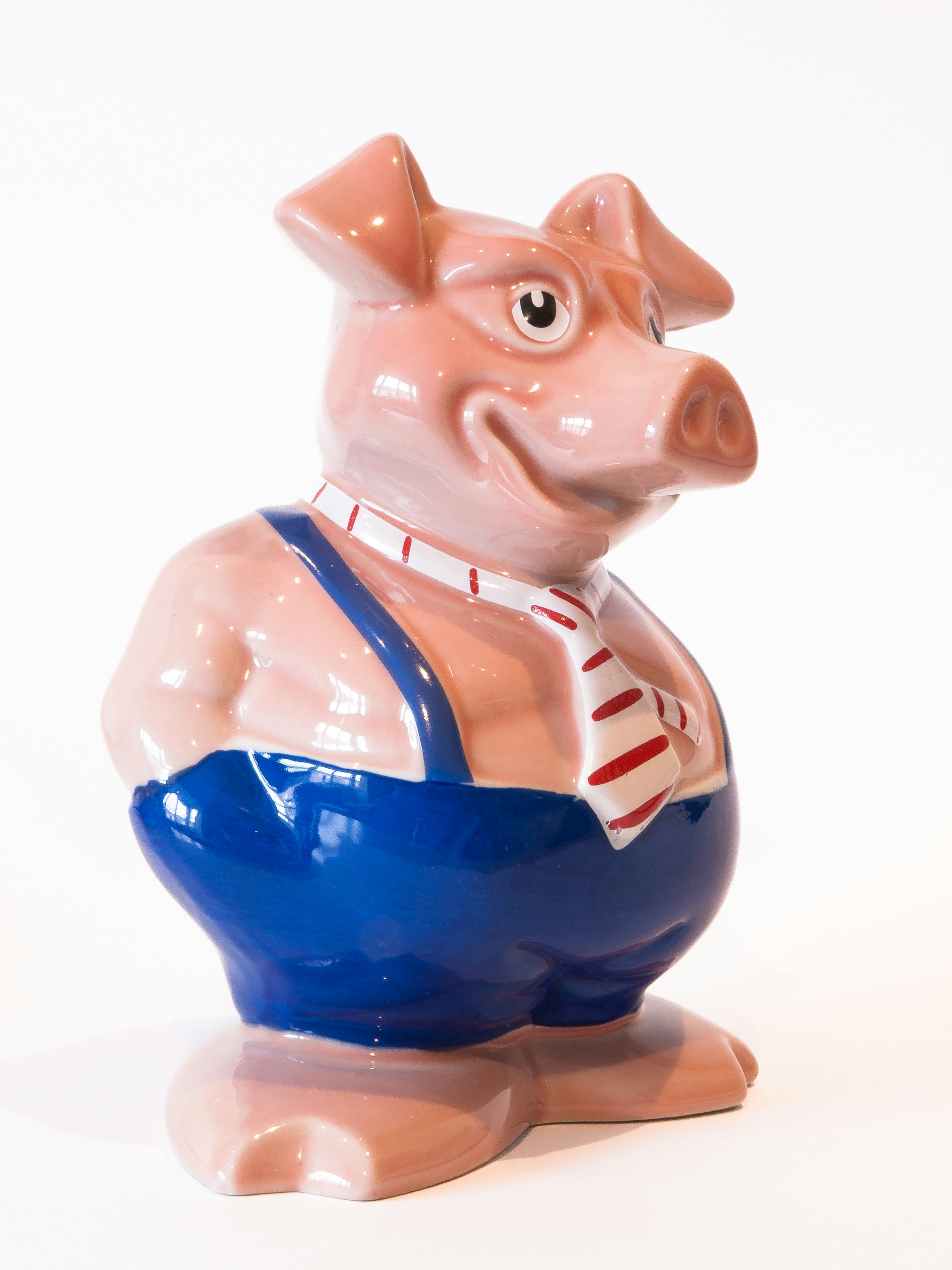 A glossy ceramic pig figurine stands on a white backdrop, dressed in blue overalls and a striped tie. This playful sculpture emphasizes glaze and character.