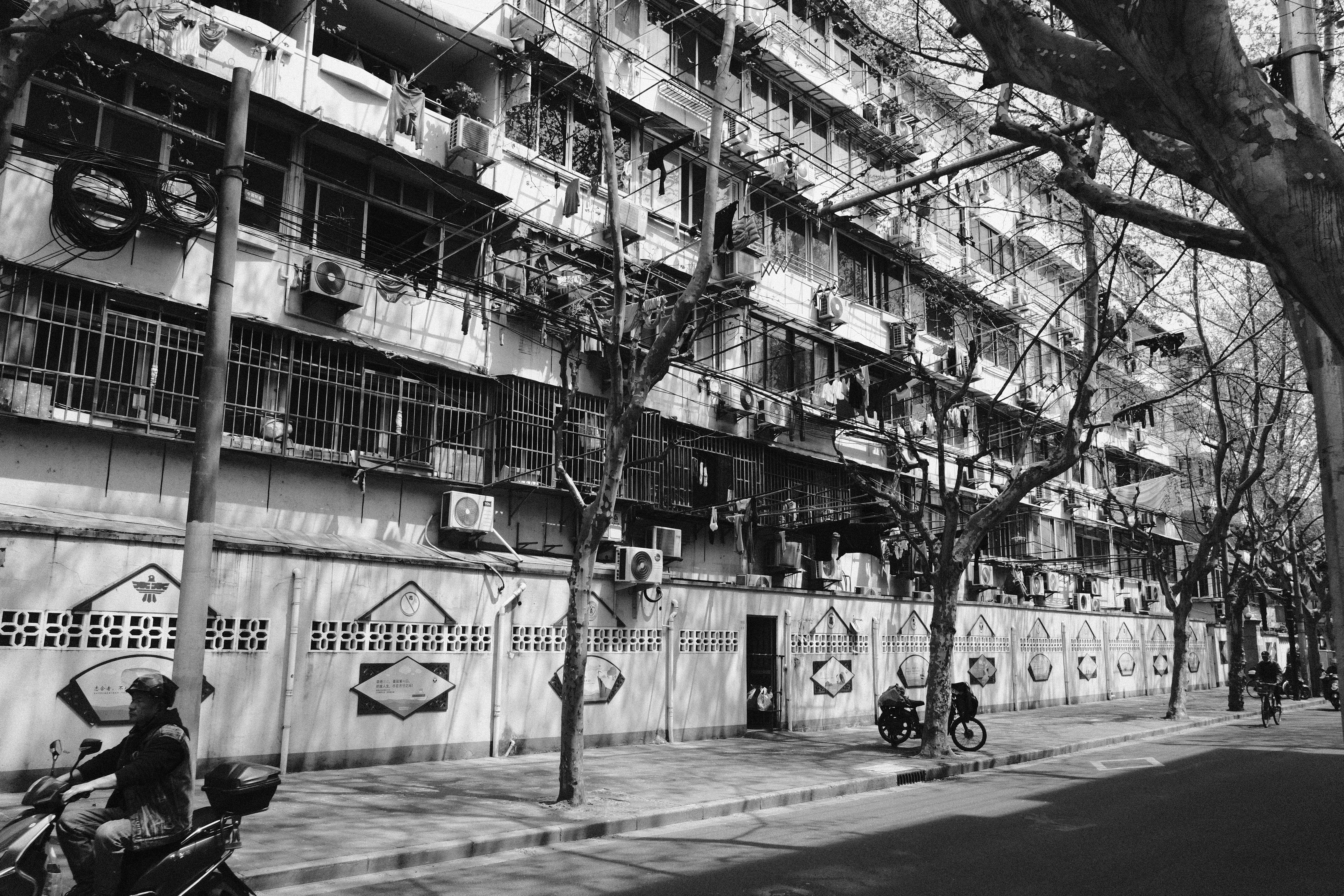 Black and white street scene with a person riding a motorcycle past a residential building lined with balconies and trees.