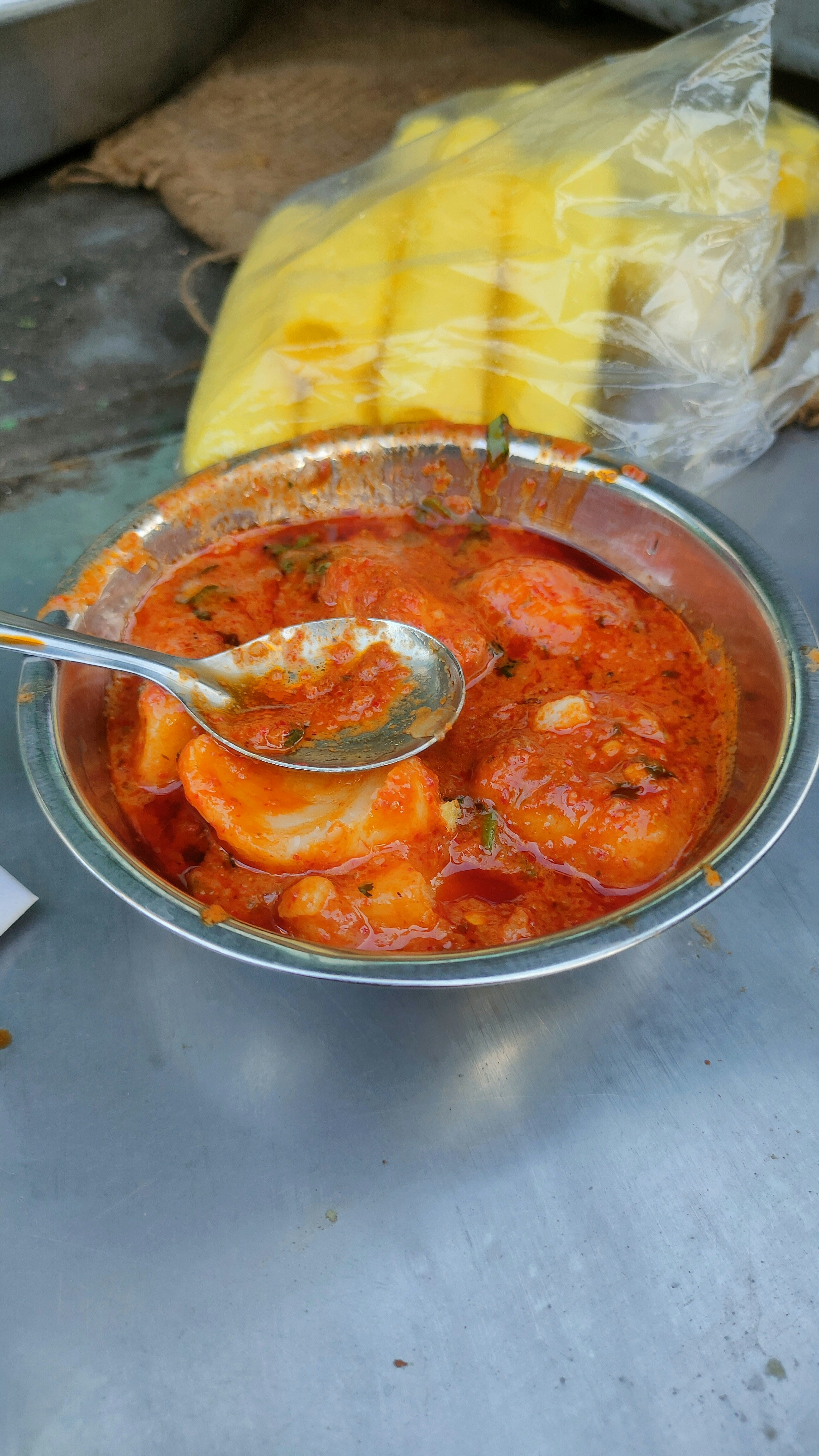 Metal bowl of red curry with a spoon on a stainless table, with a street-food setup in the background.