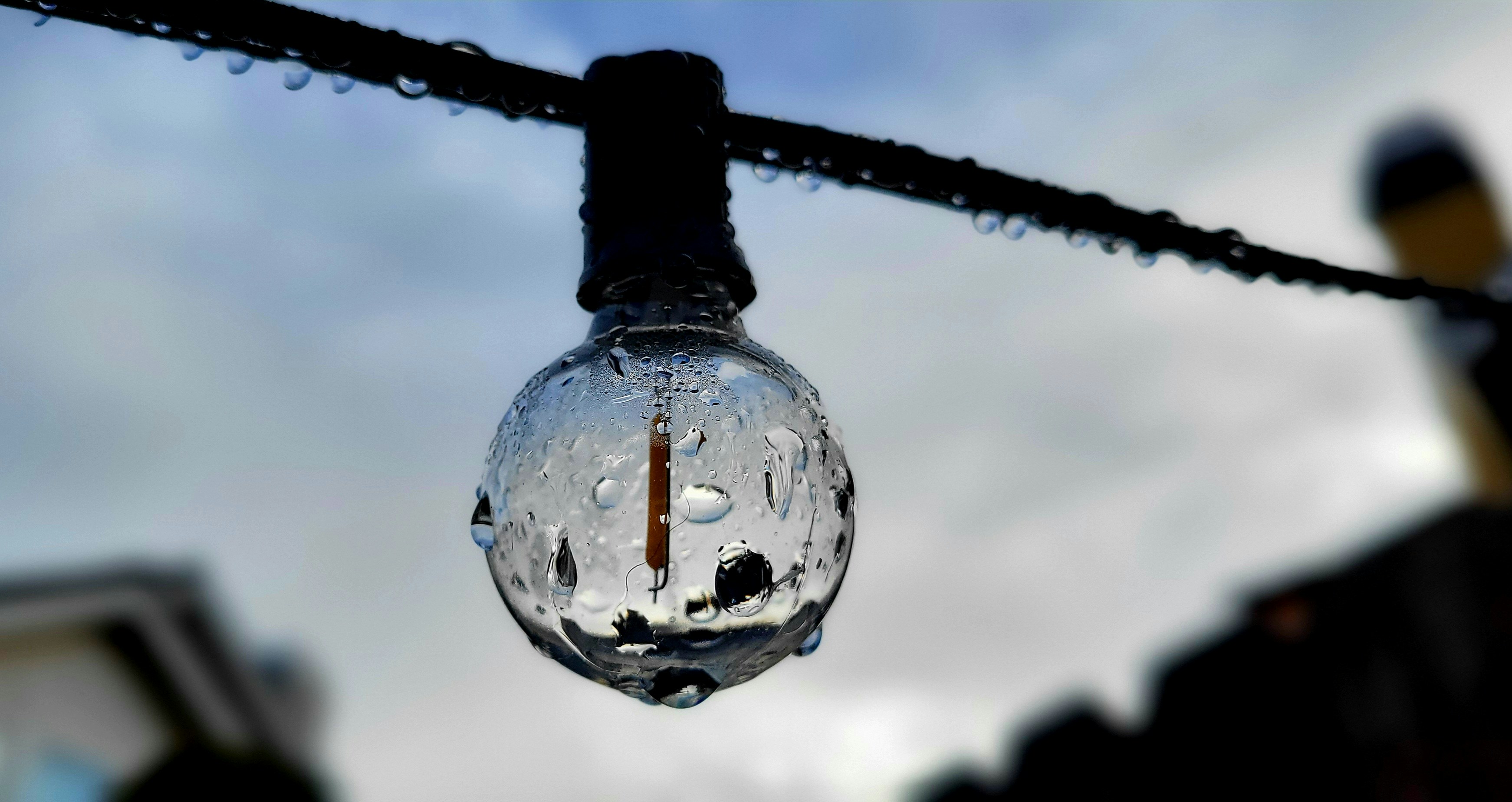 A single glass water droplet hangs from a wire, acting as a lens with a softly blurred background. This close-up photograph emphasizes refraction, texture, and the subtle glow on wet glass.