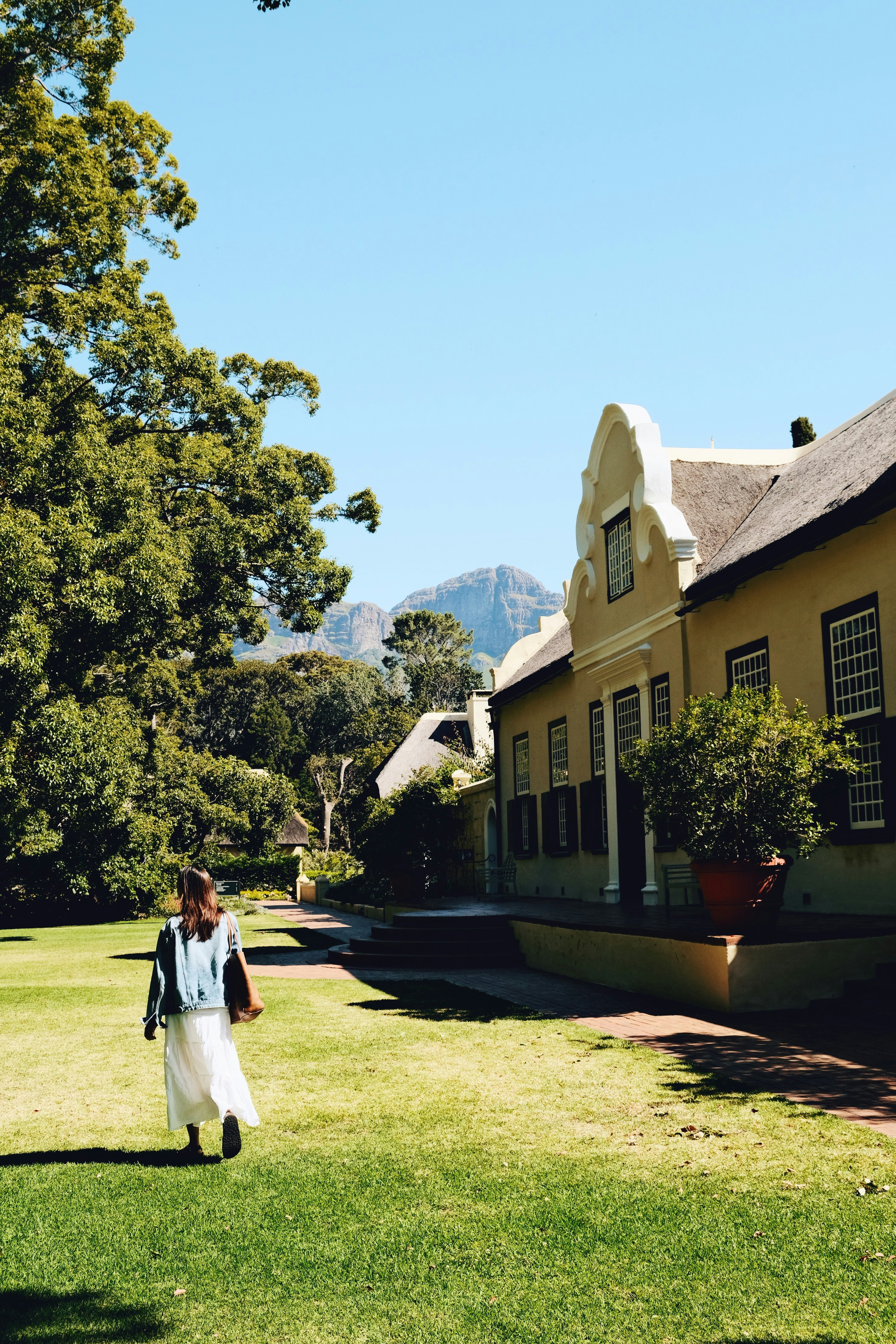 Sunlit Threshold: A lone traveler on a grassy path toward a yellow colonial-style building
