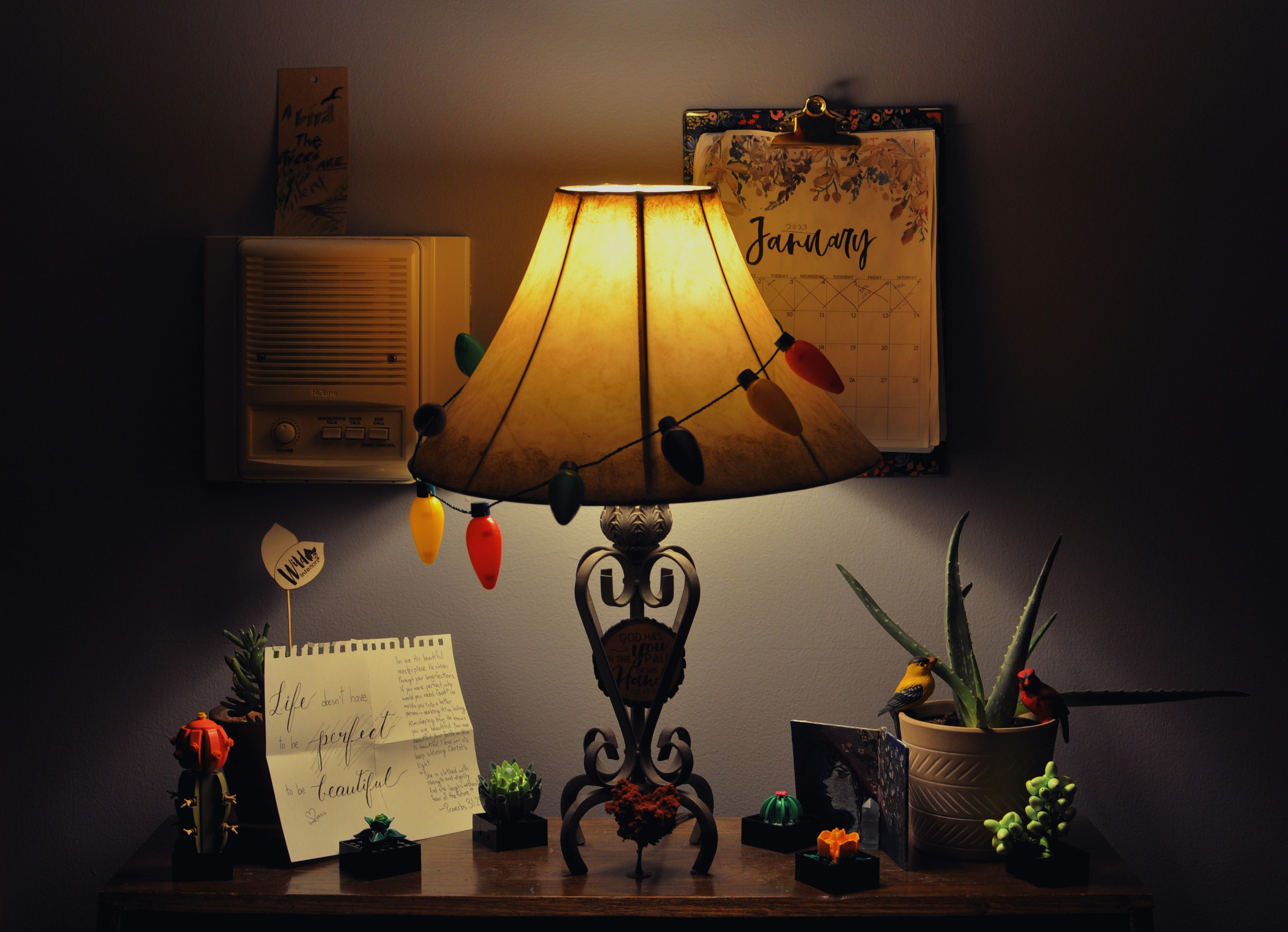 A vintage lamp adorned with colorful lights illuminates a cozy table setting featuring plants, notes, and a calendar. The soft light creates a welcoming atmosphere.