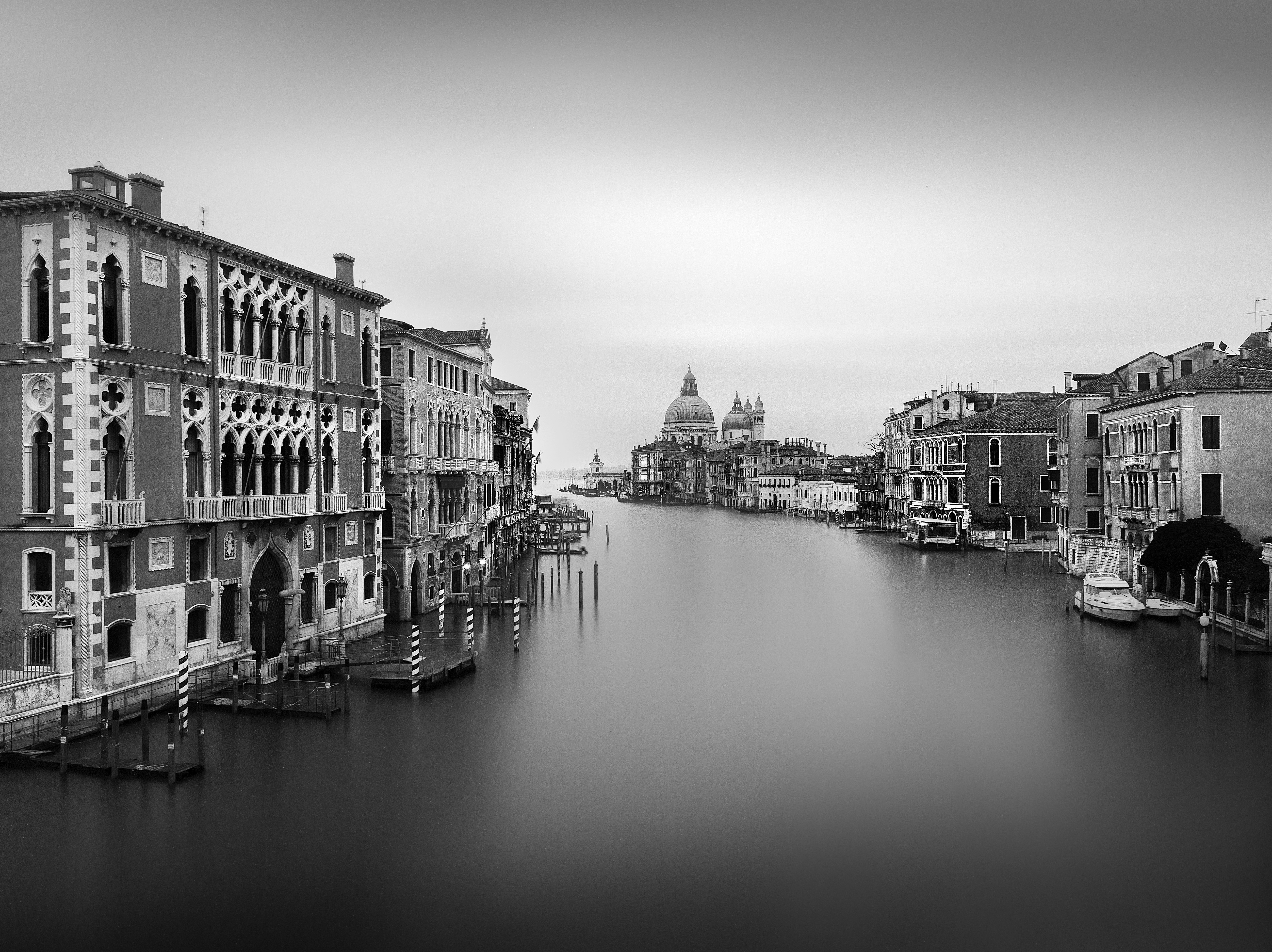 This captivating black and white image captures the timeless allure of Venice's Grand Canal from the Ponte dell'Accademia. The smooth, ethereal water contrasts with the ornate facades of historic buildings, leading the eye towards the majestic dome of Santa Maria della Salute in the distance. The soft, diffused lighting enhances the tranquil atmosphere, creating a visually striking composition that evokes a sense of calm and nostalgia.