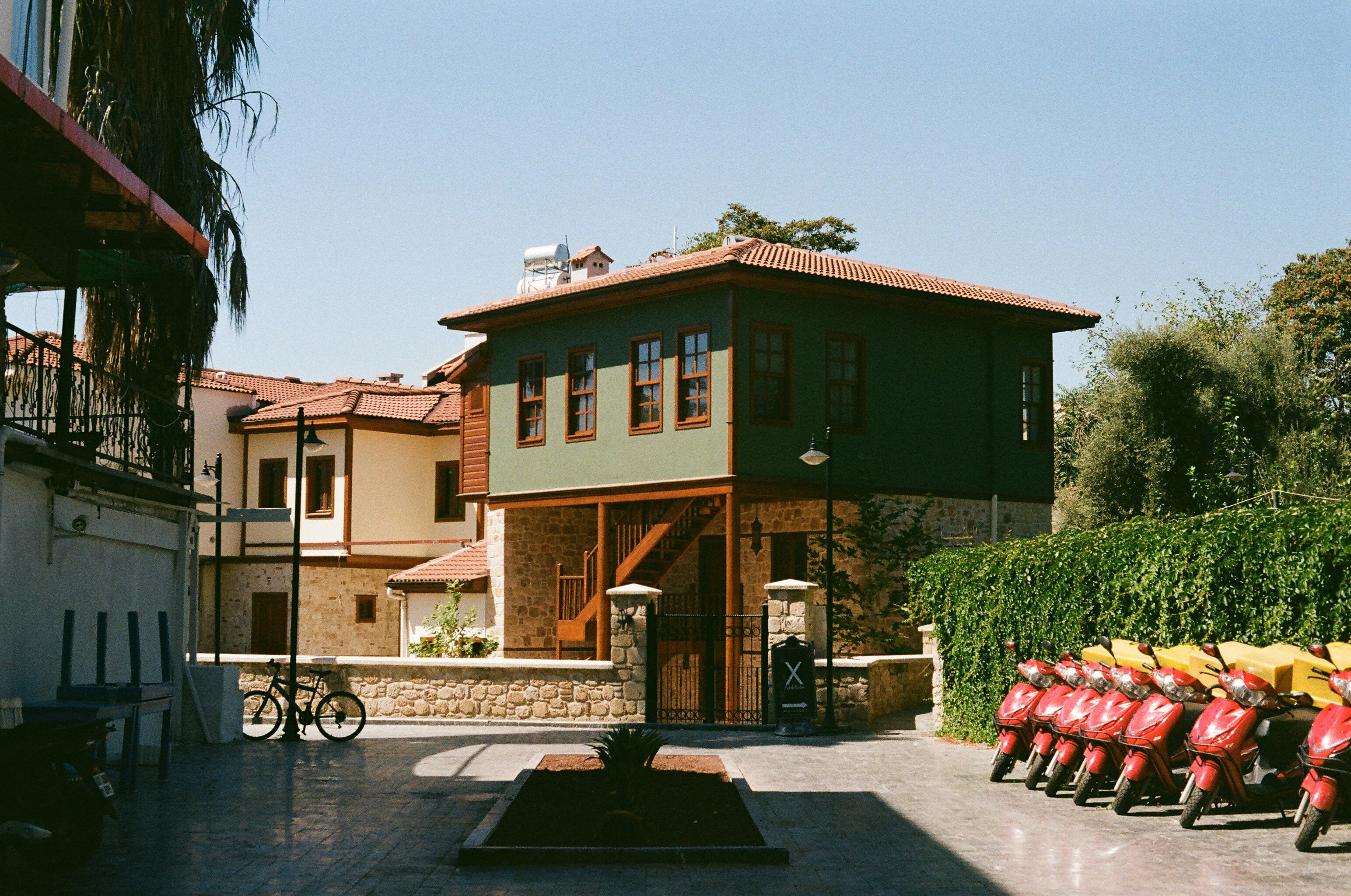 Sunlit street scene photograph of a two-story green house with brown trim in a courtyard, with a line of red scooters on the right.