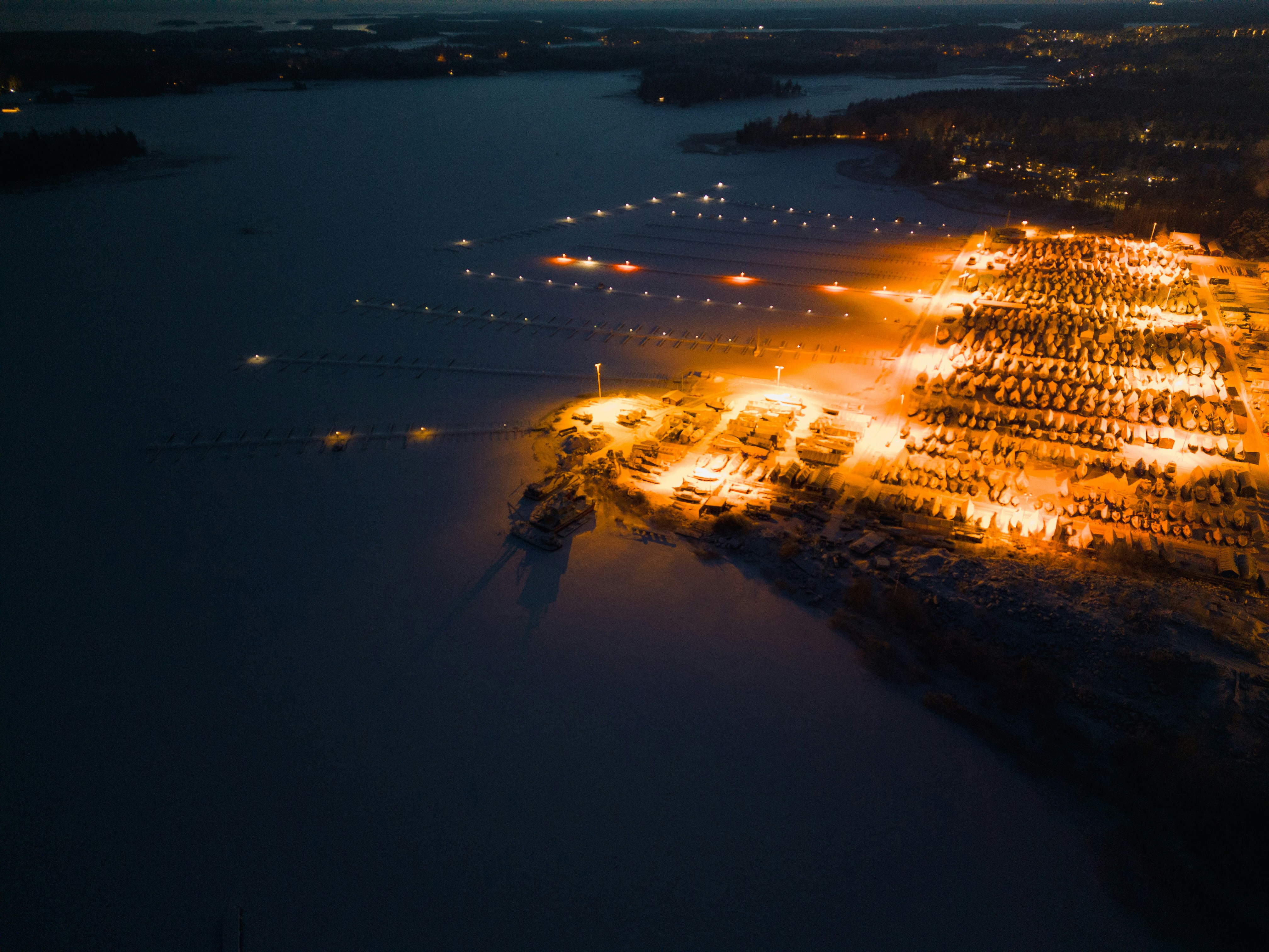 Nighttime drone photograph of a coastal marina lit by rows of warm lights along the shore. Calm water mirrors the glow while a grid of berths and piers defines the illuminated coastline.