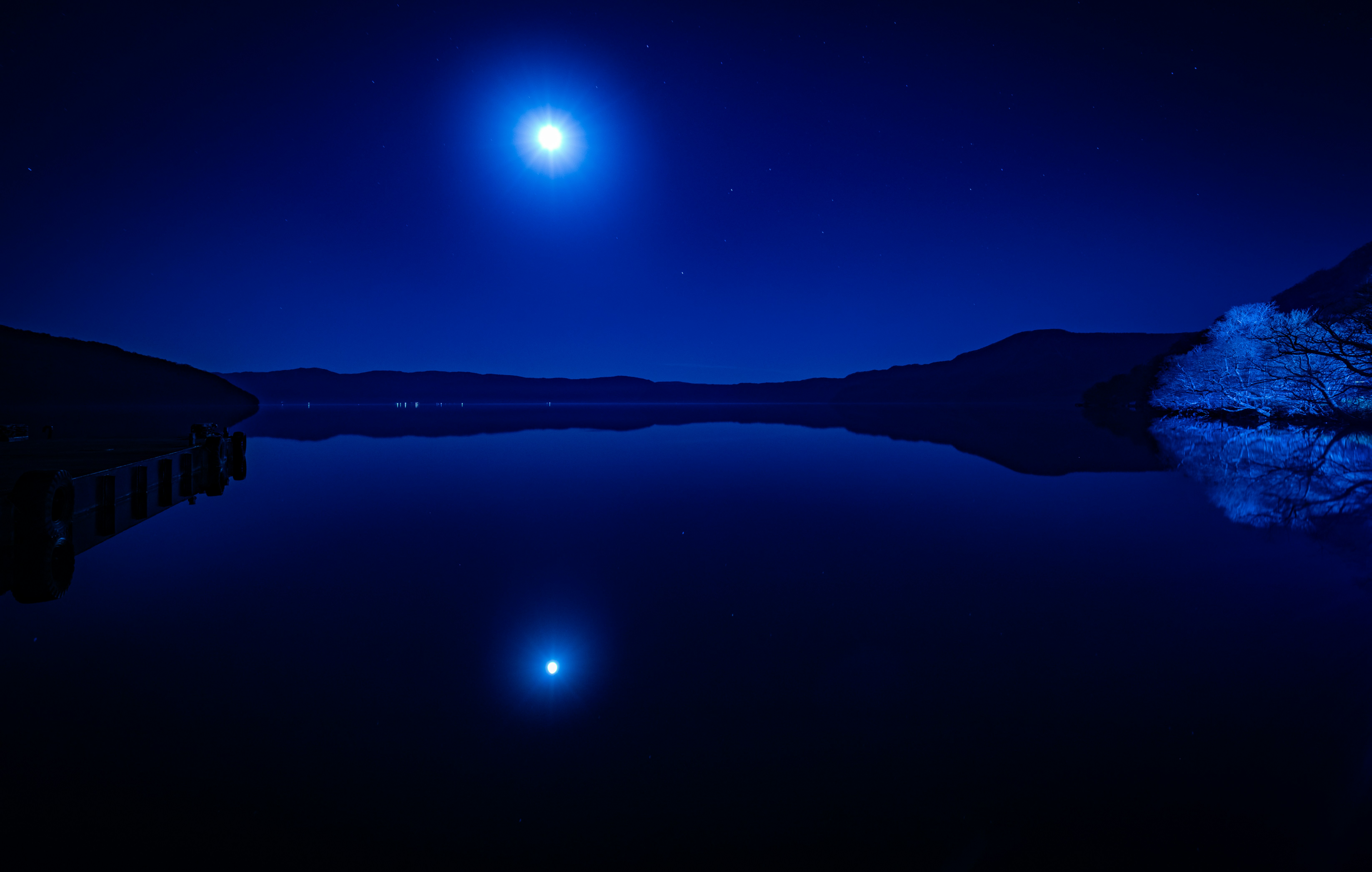 Bright full moon reflecting on a calm lake under a deep blue night sky.