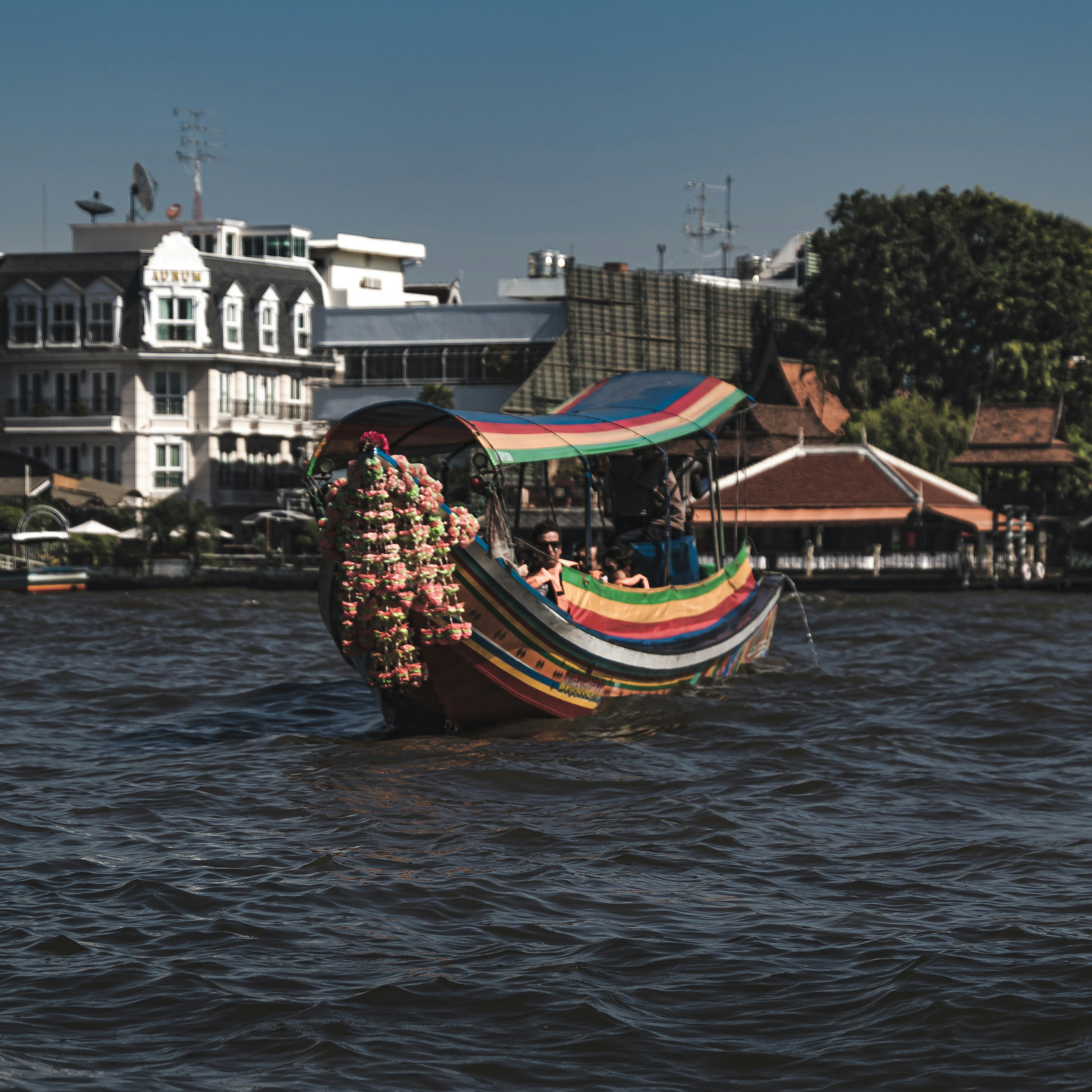 Garland-Adorned Long-Tail Boat Gliding Past a Riverside Village