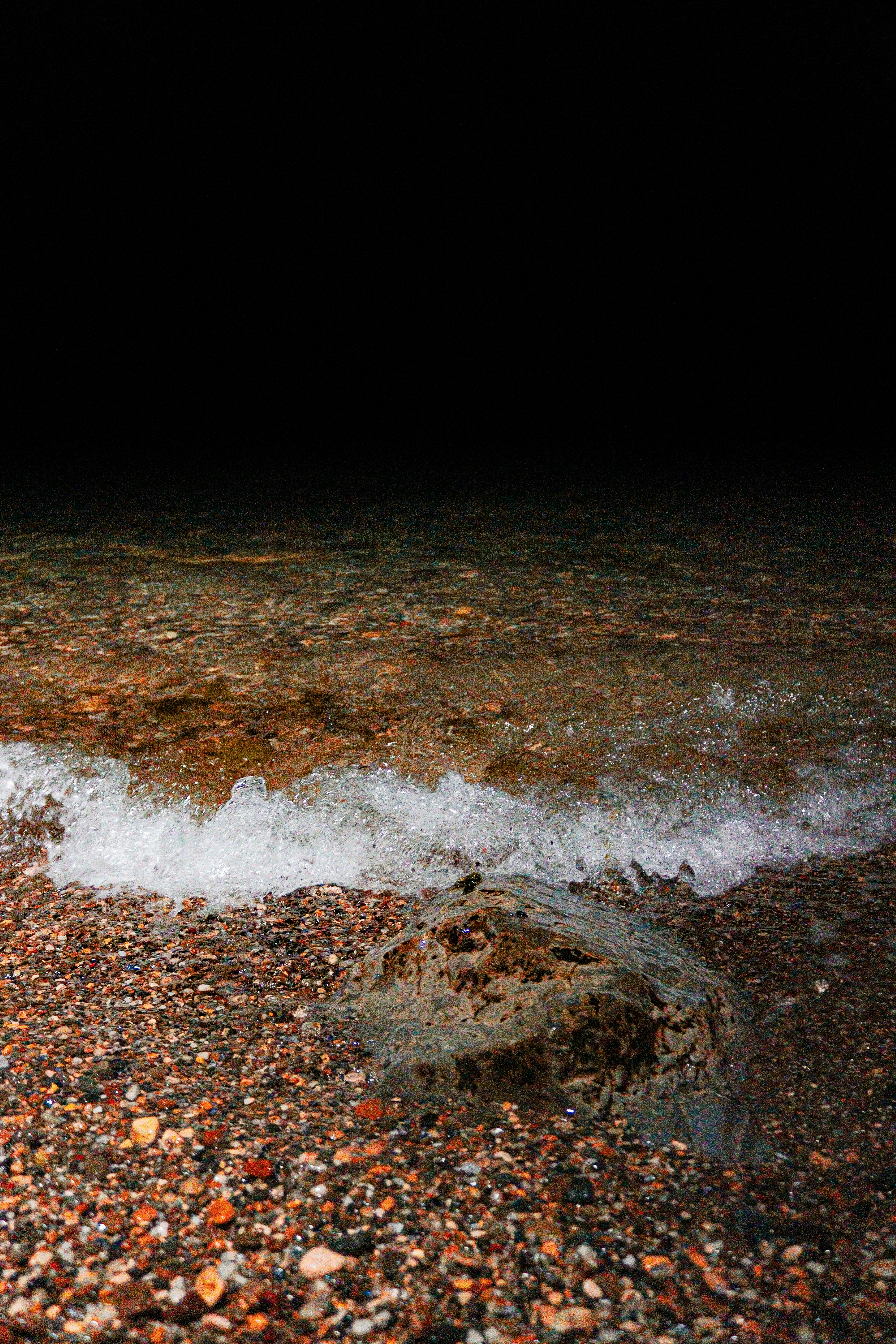 Gentle waves lap against a rocky beach, illuminated by a soft glow in the darkness. The water reflects the subtle hues of the night sky.