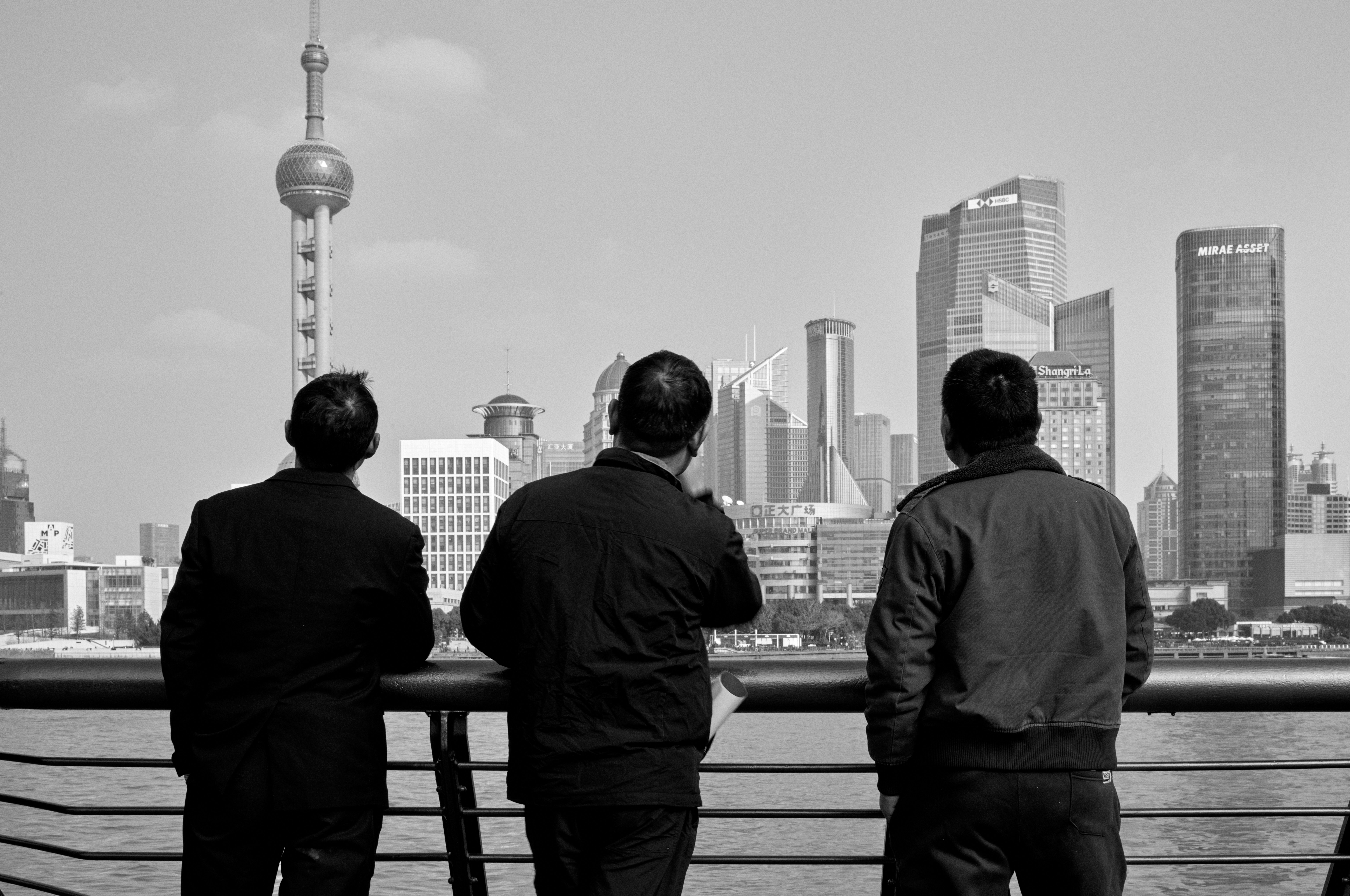Three figures in dark jackets stand with their backs to the viewer, facing a city skyline across a river; a railing separates them from the water, creating a contemplative urban scene.