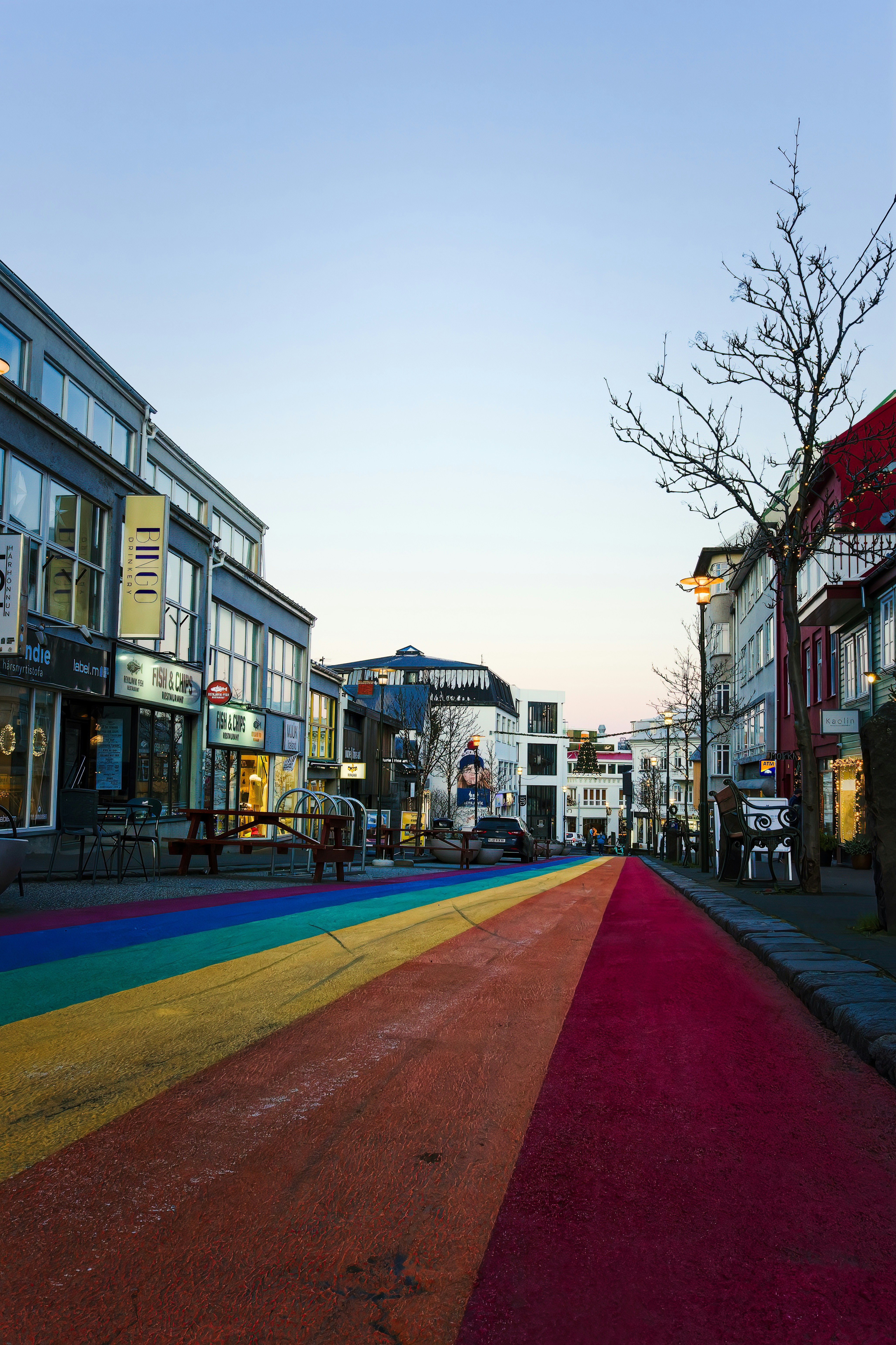 This captivating image captures a vibrant rainbow crosswalk stretching down a bustling street in Reykjavik, Iceland. Flanked by colorful buildings on either side, the scene is set against the backdrop of a serene pale blue sky at dusk, with streetlights gently illuminating the path. The contrast between the vivid crosswalk and the soft evening light creates a visually striking and inviting atmosphere.