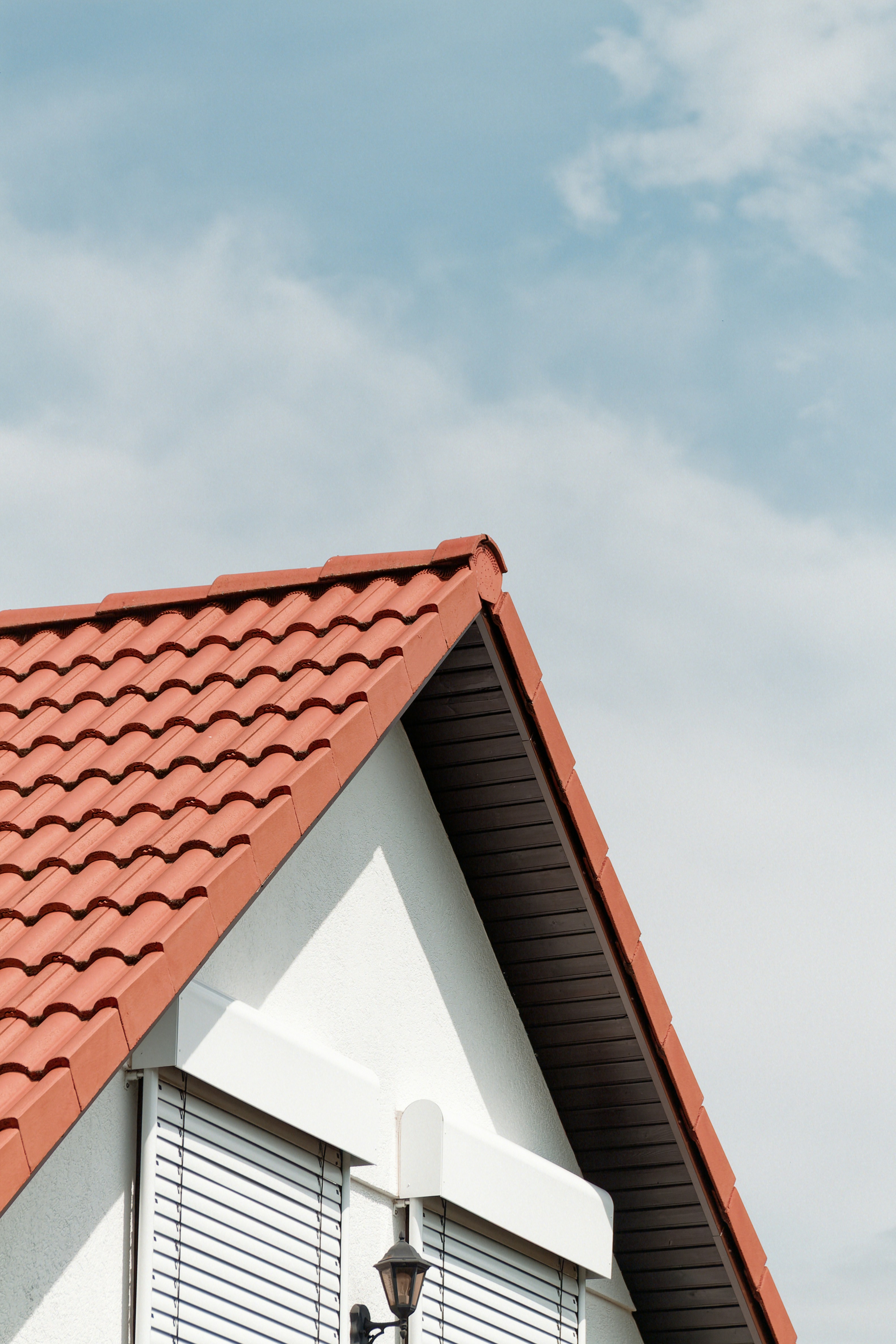A close-up view of a house's roof featuring terracotta tiles and a decorative lamp, framed against a soft, cloudy sky.