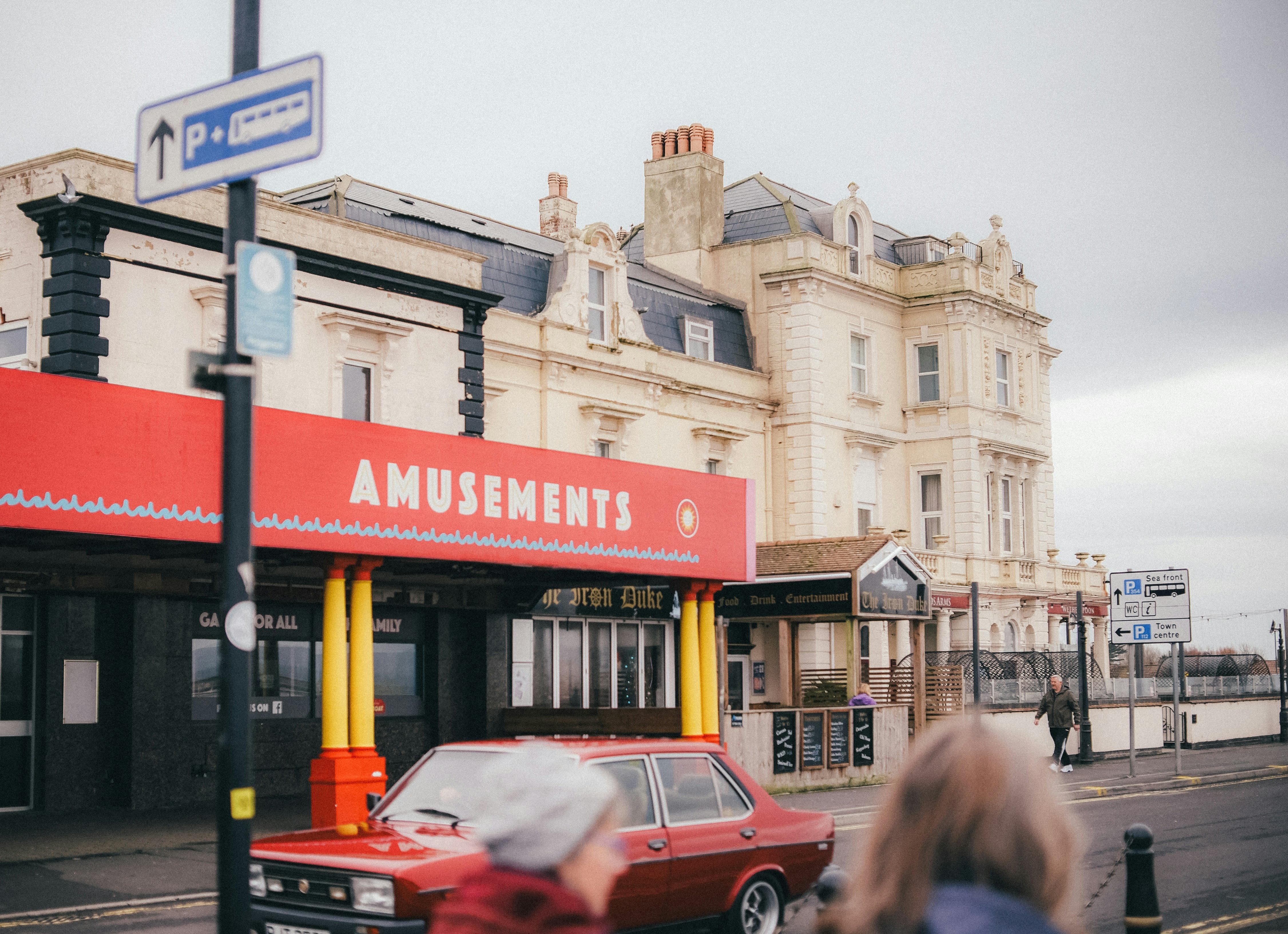 This image captures a vibrant seaside amusement area with a striking red and yellow awning that contrasts against the soft, overcast sky. The composition includes a vintage car and classic architecture, evoking a sense of nostalgia. The muted tones and diffused lighting create a calm and inviting atmosphere, making the scene visually compelling.