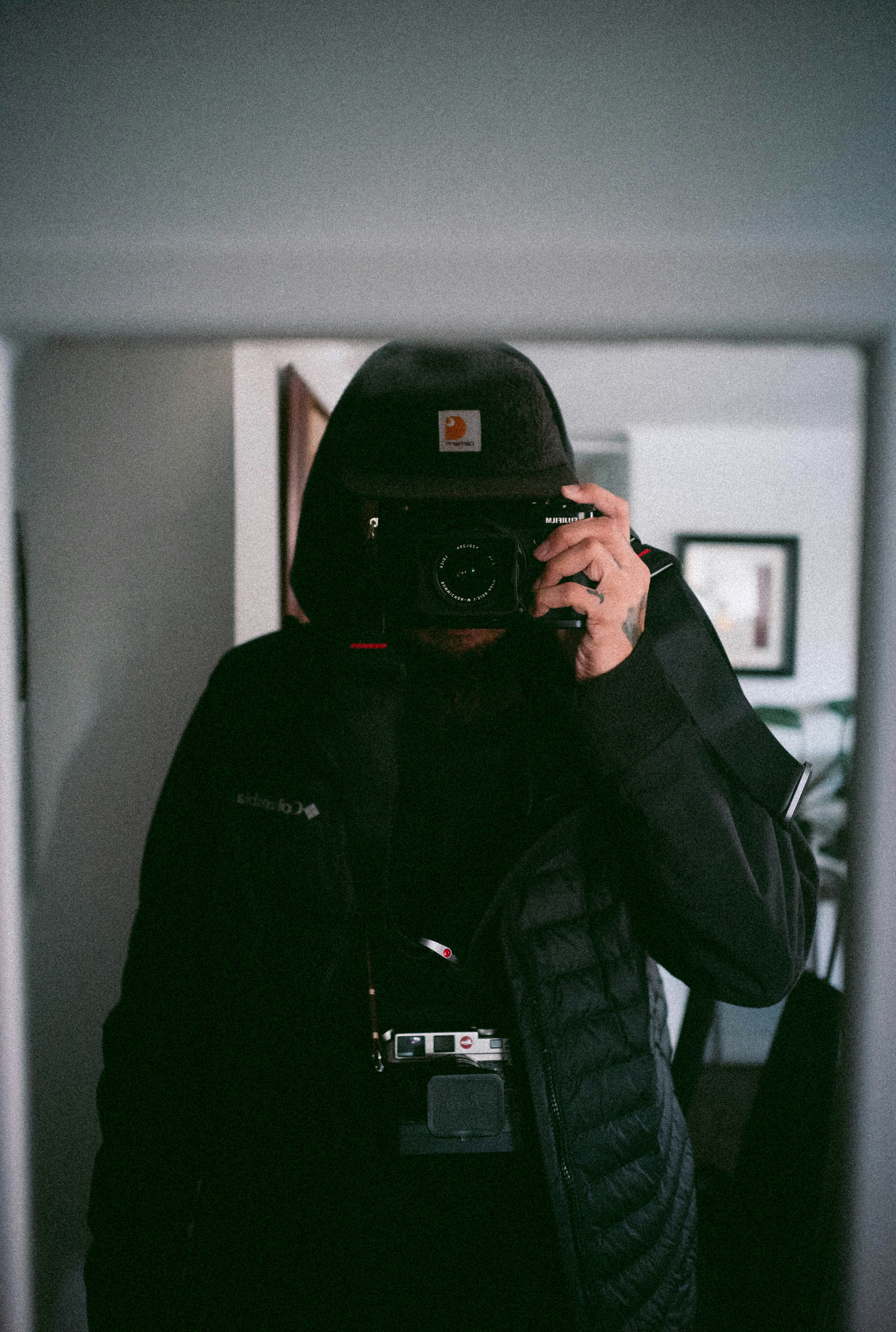 Dim indoor mirror photograph of a hooded photographer holding a camera.