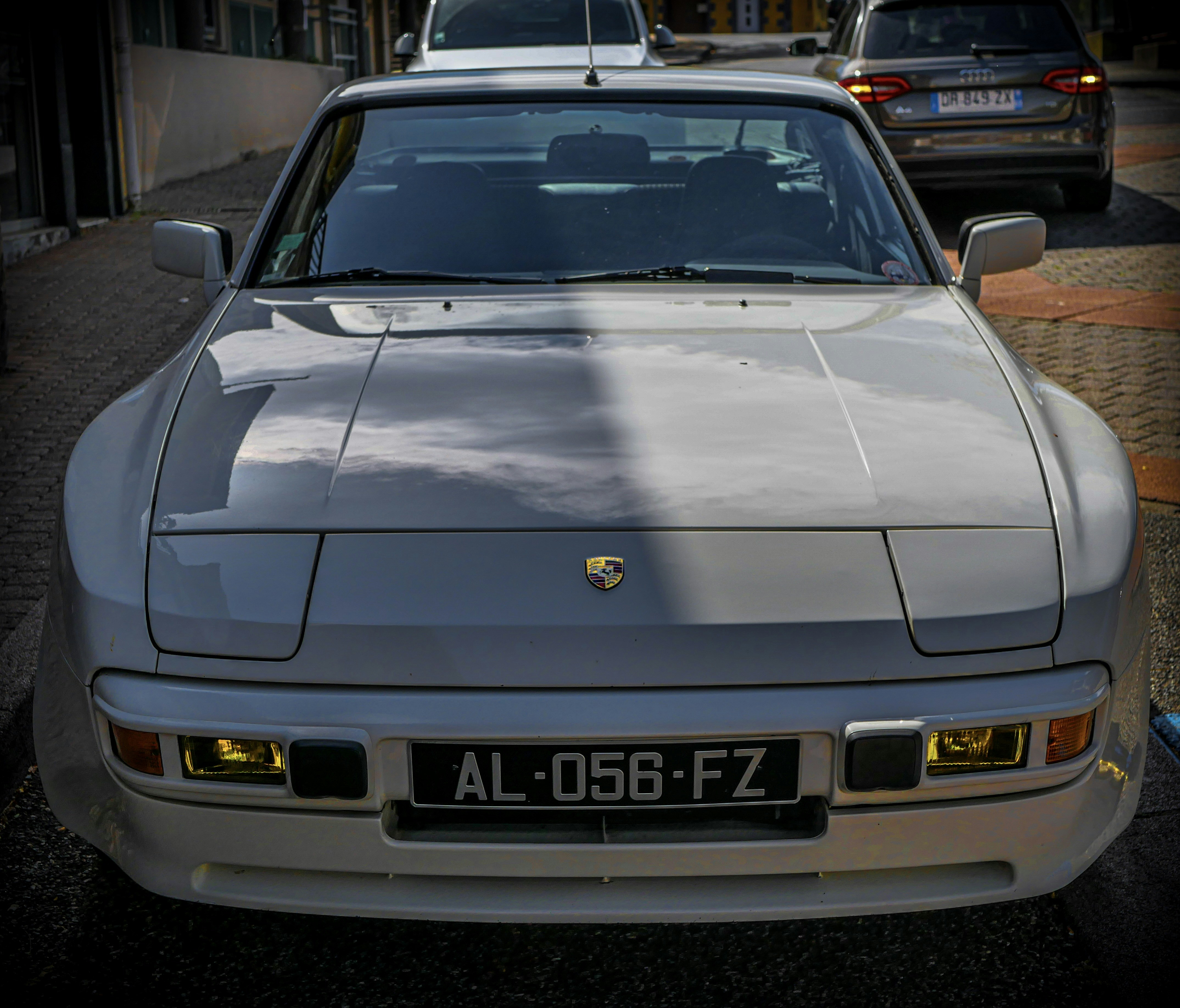 White Porsche with cloud reflection on the hood, parked on a city street.