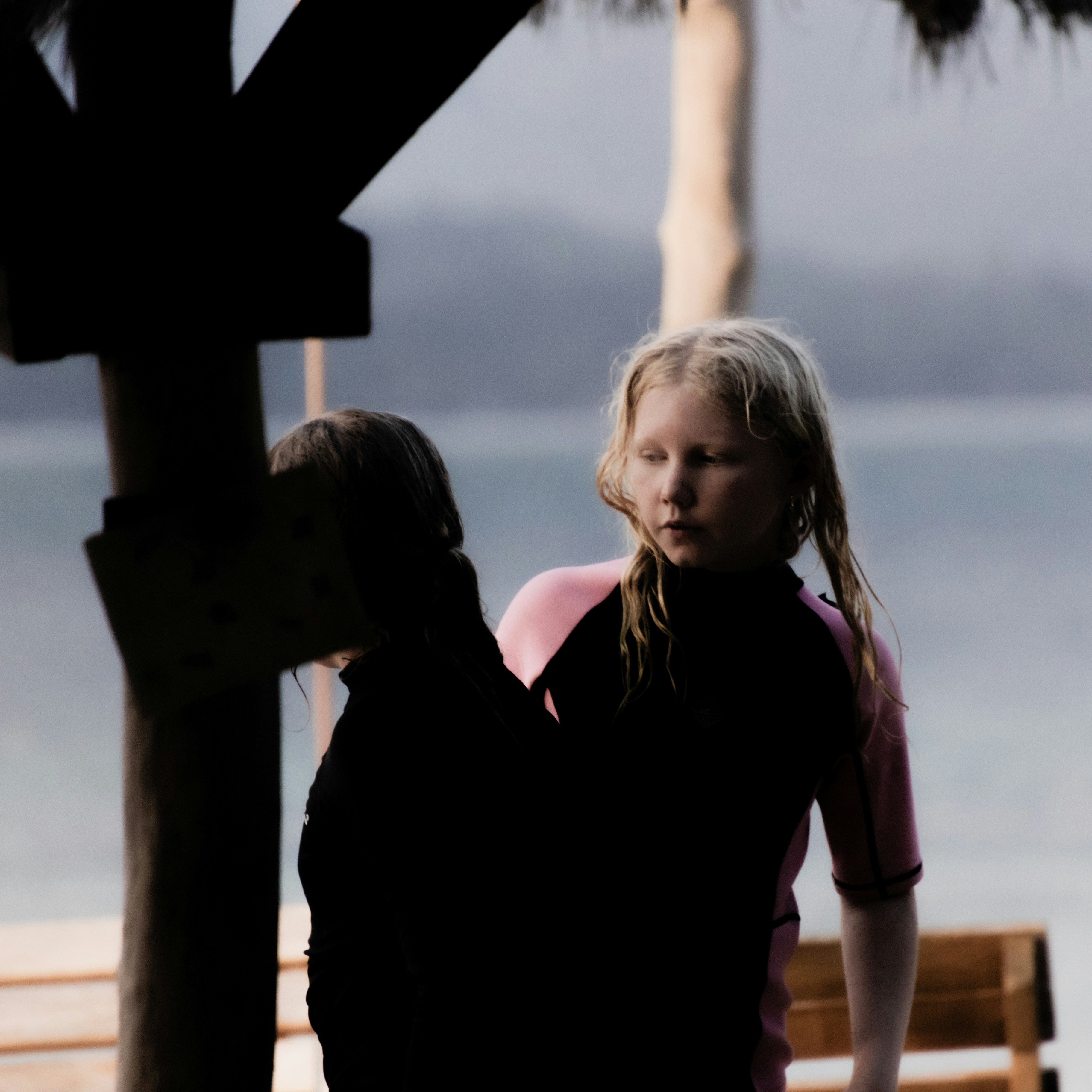 Two children share a quiet moment under a shaded structure near the water, capturing the innocence of youth. Their expressions reflect a serene connection with each other and their surroundings.