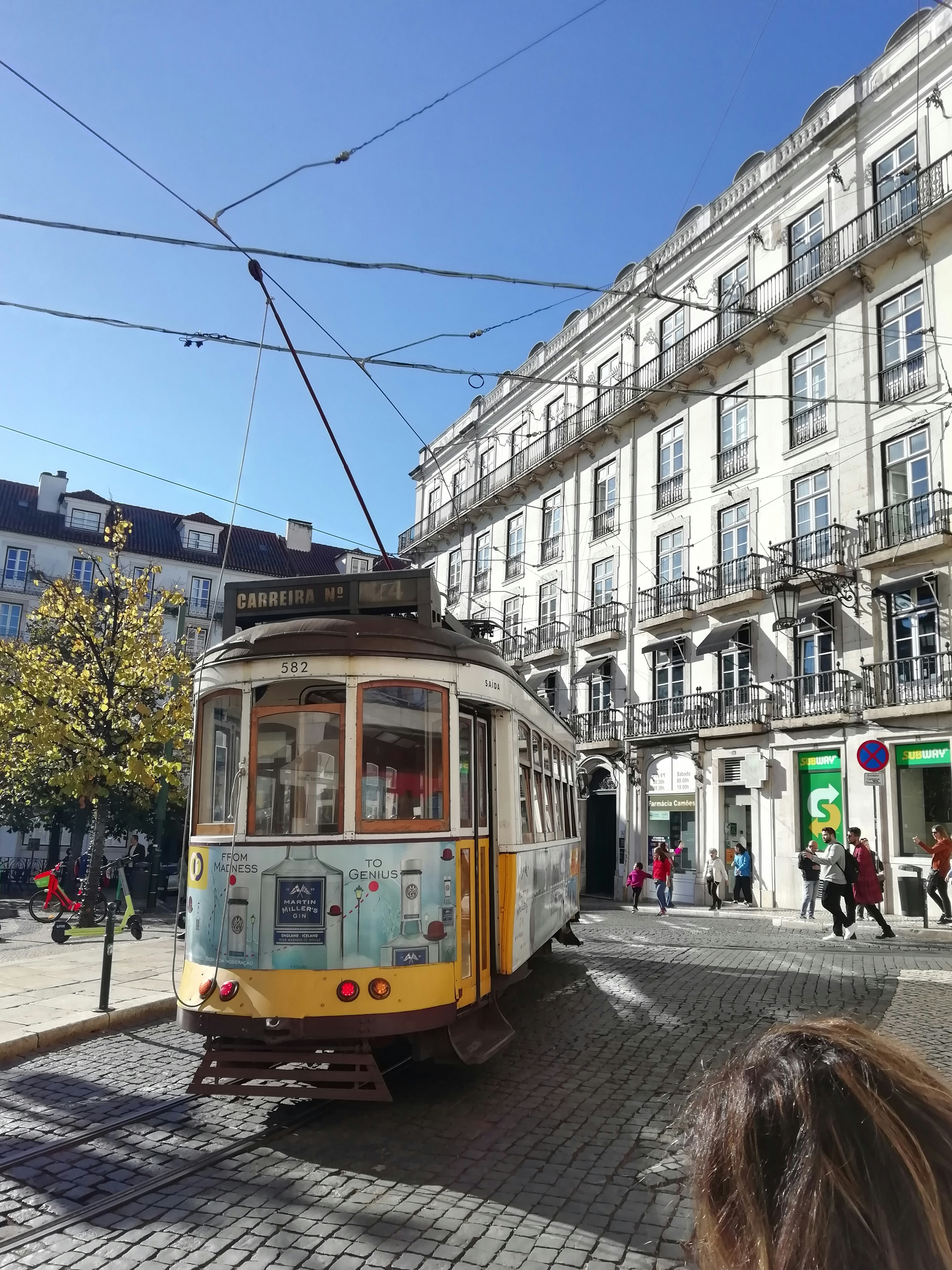 A vintage tram pauses on cobblestones beside a white, columned building. Pedestrians move along the square as overhead wires trace the bright blue sky.