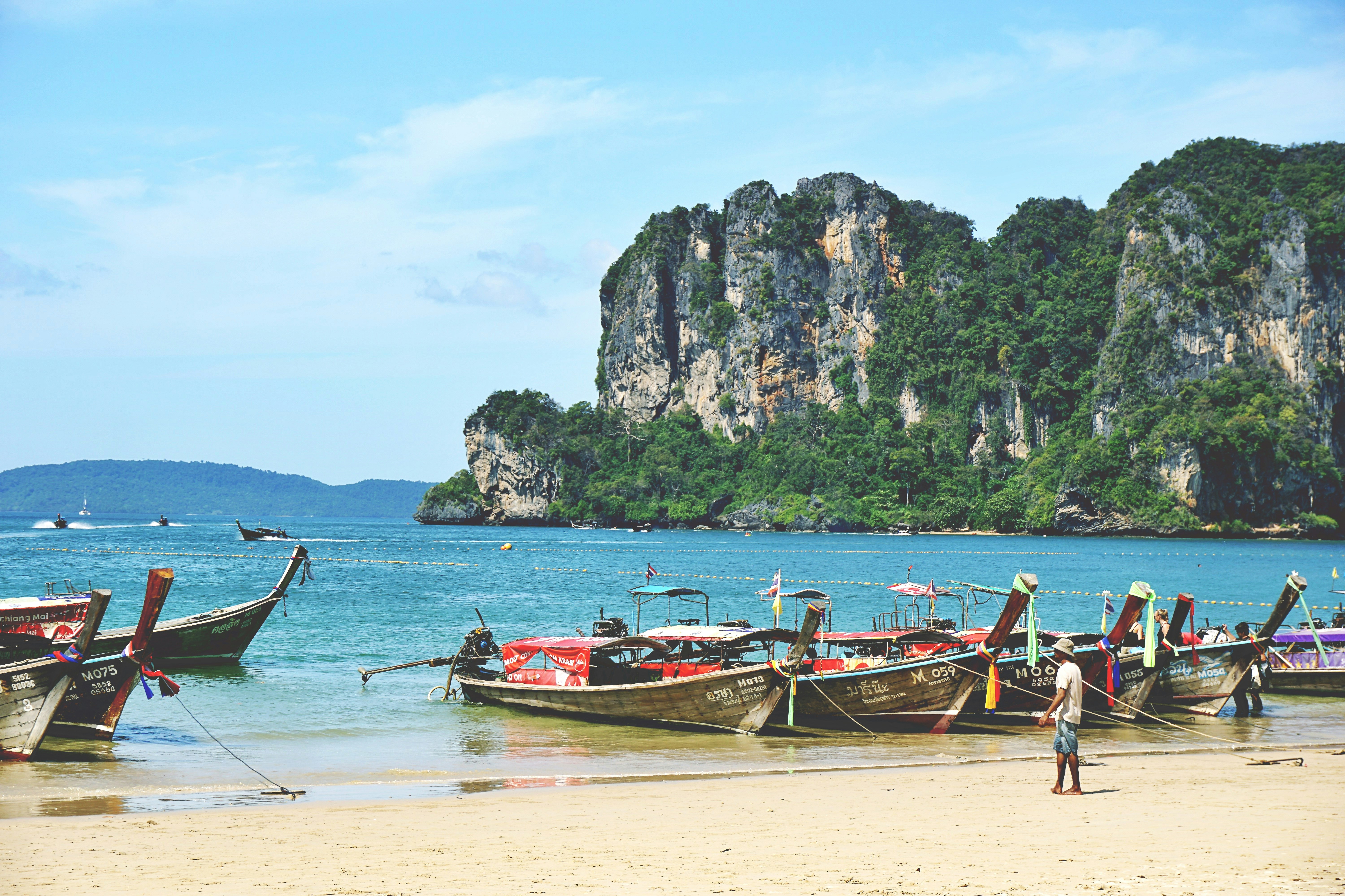 Fishing boats anchored on a tranquil beach with lush cliffs in the background. A lone figure strolls along the shoreline.
