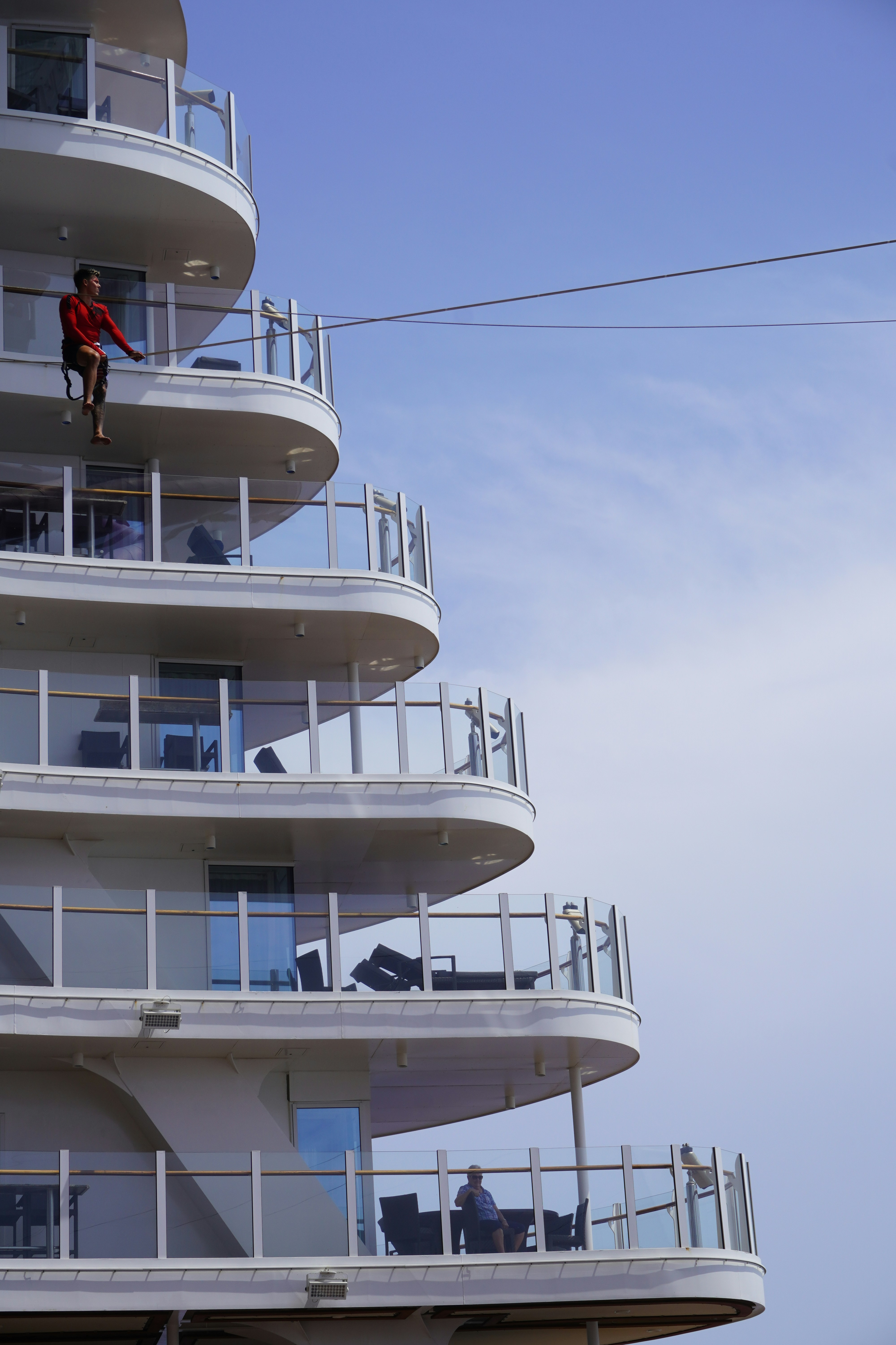 A worker in a red shirt is suspended on a cable, performing maintenance on a modern building with multiple balconies. The scene captures the tension between human endeavor and architectural design.