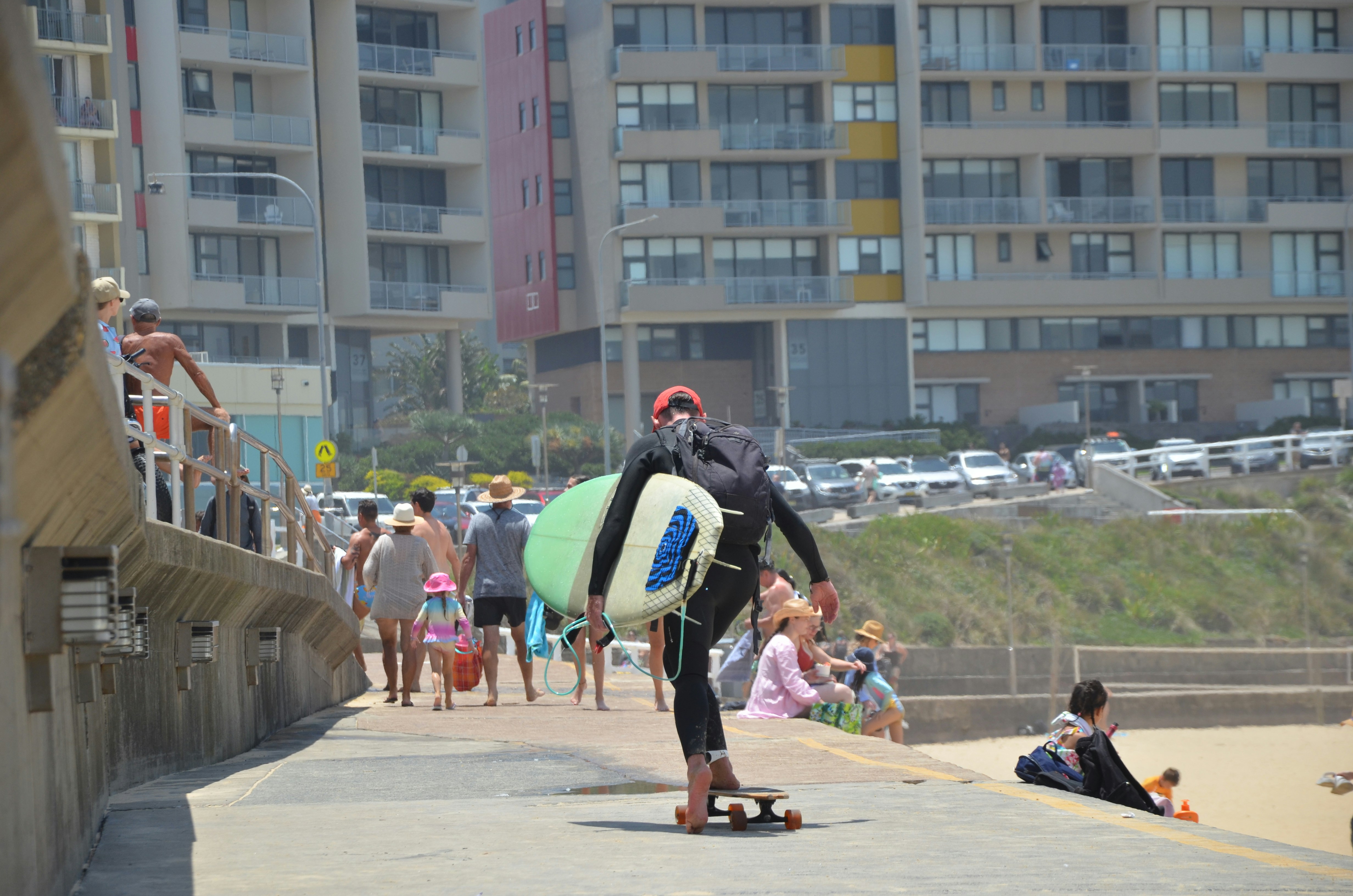 Surfer on skateboard heading towards the beach, surfboard in tow, amidst a lively crowd enjoying the sun. 