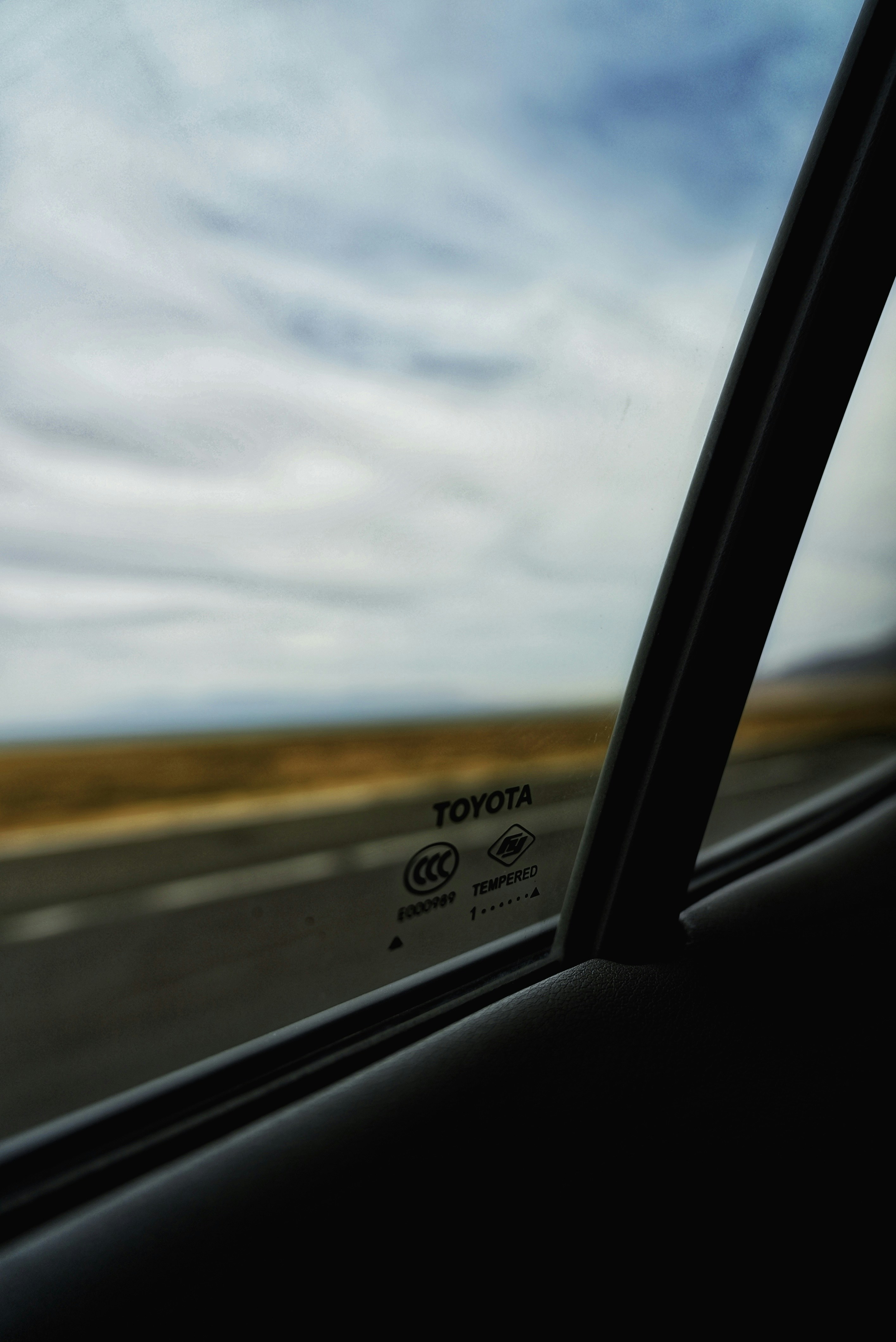 A photograph from inside a car shows a Toyota logo decal on the glass, with a blurred highway and blue sky beyond. The shallow depth of field emphasizes the decal while keeping the exterior scenery soft in the background.