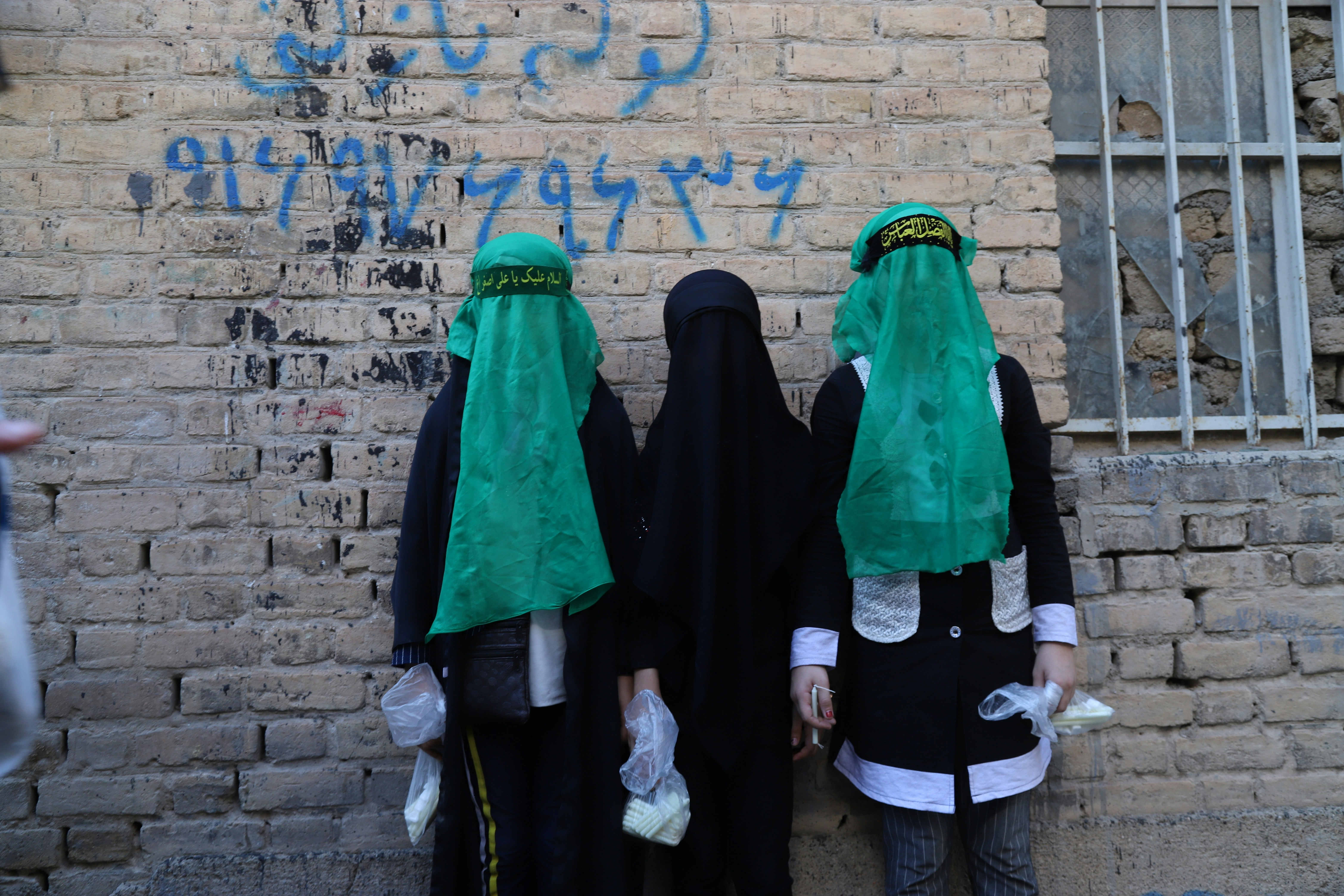 Three individuals stand in traditional attire with green and black garments against a graffiti-covered brick wall.